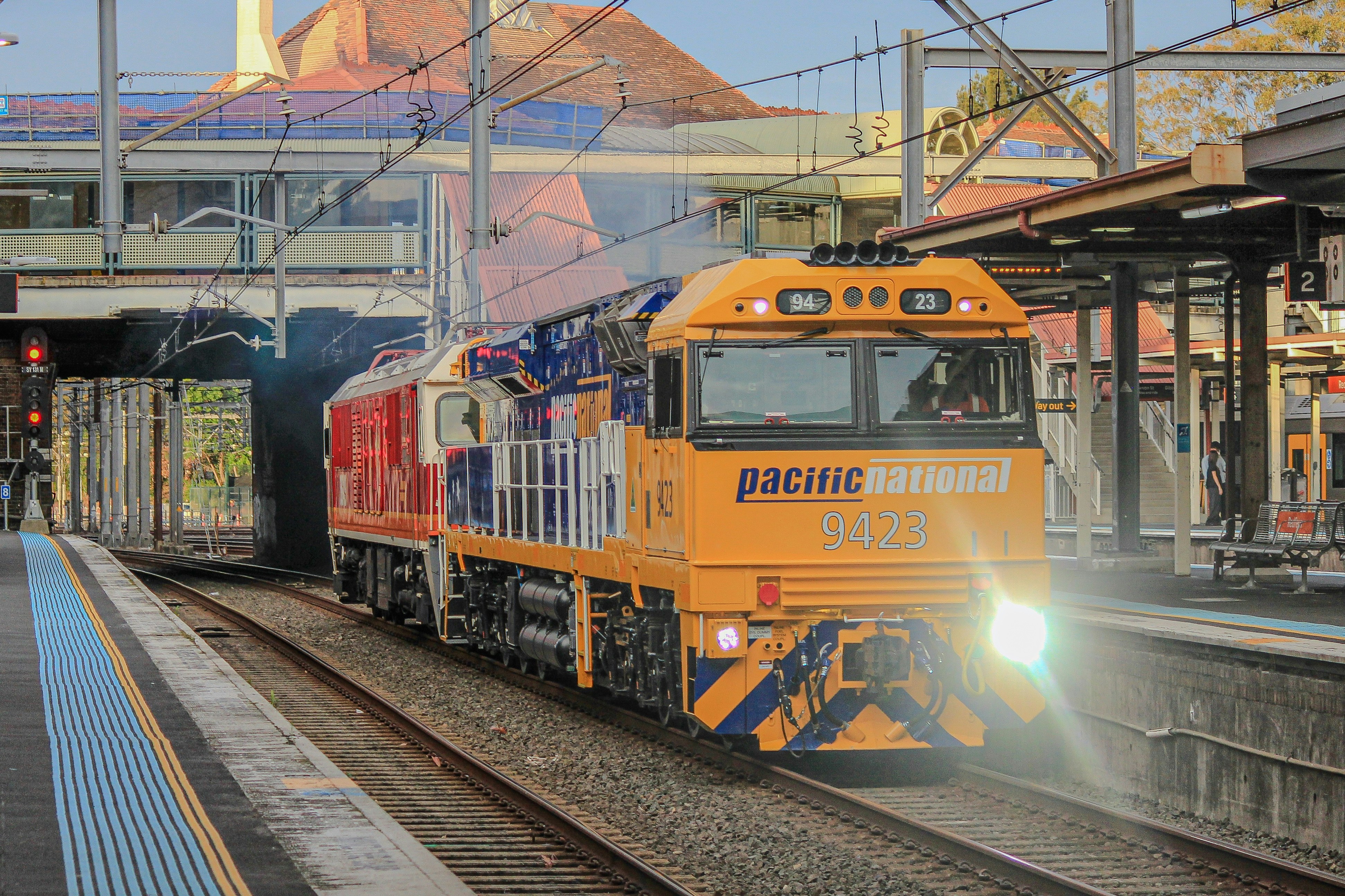 Bright orange Pacific National locomotive powers through a station, headlights blazing as passengers and tracks unfold along the platform.