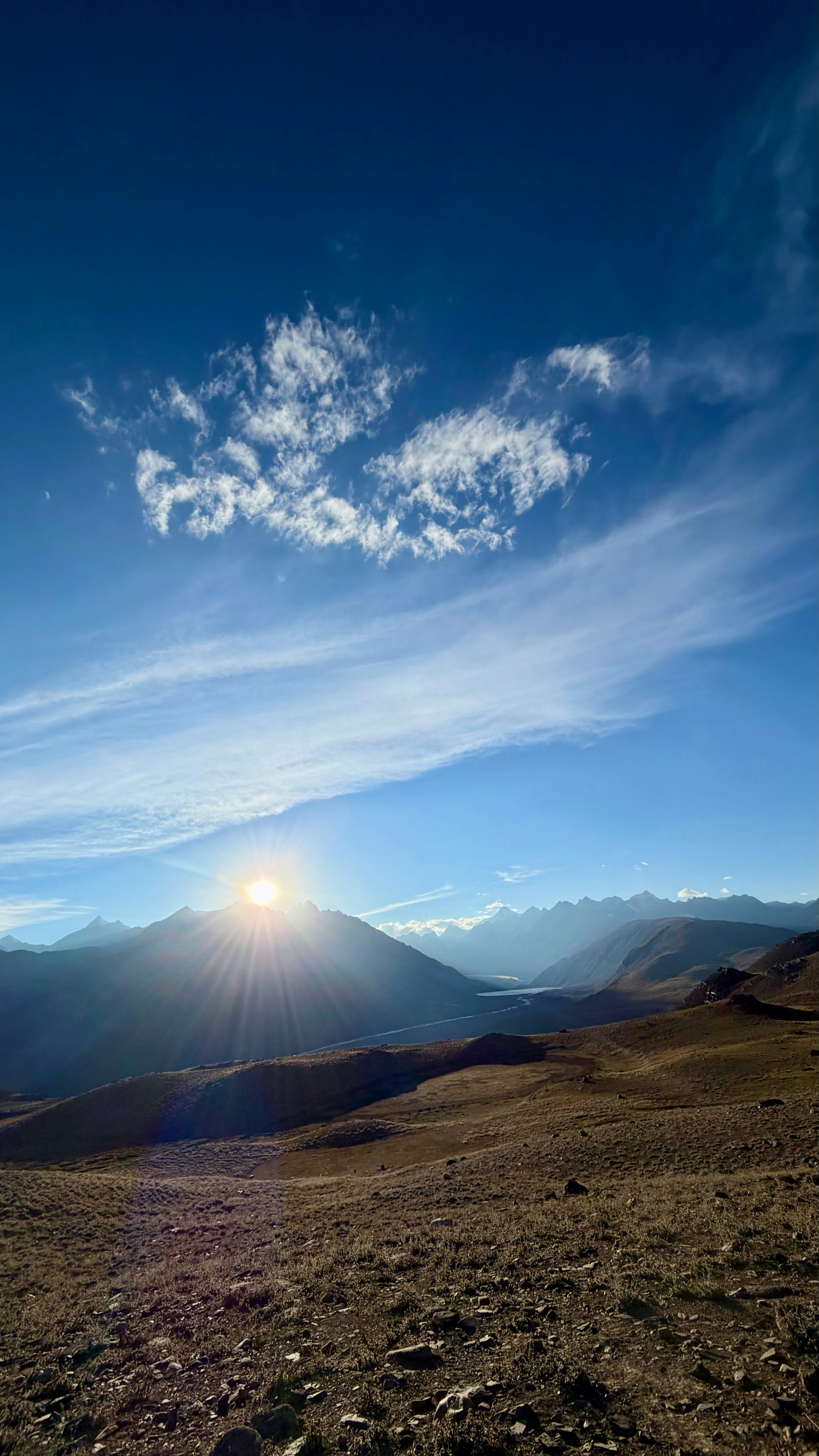 Dramatic mountain landscape at sunrise with the sun peeking over a ridge and a wide valley below; the scene emphasizes vast sky, alpine terrain, and a tranquil, expansive atmosphere. Logo-free natural landscape.