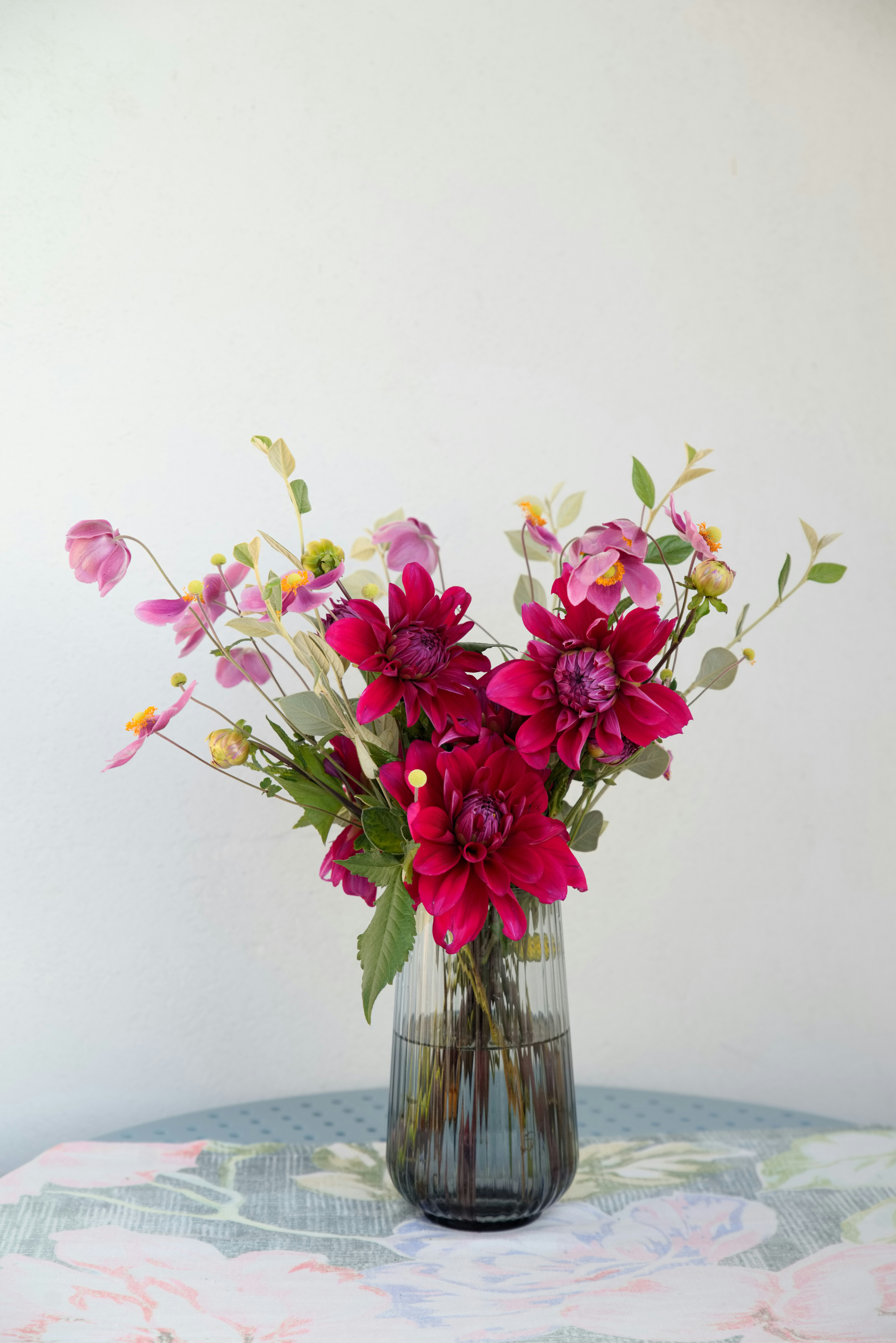 Vibrant pink bouquet gracefully arranged in a ribbed glass vase on a floral-patterned tablecloth against a pale wall.