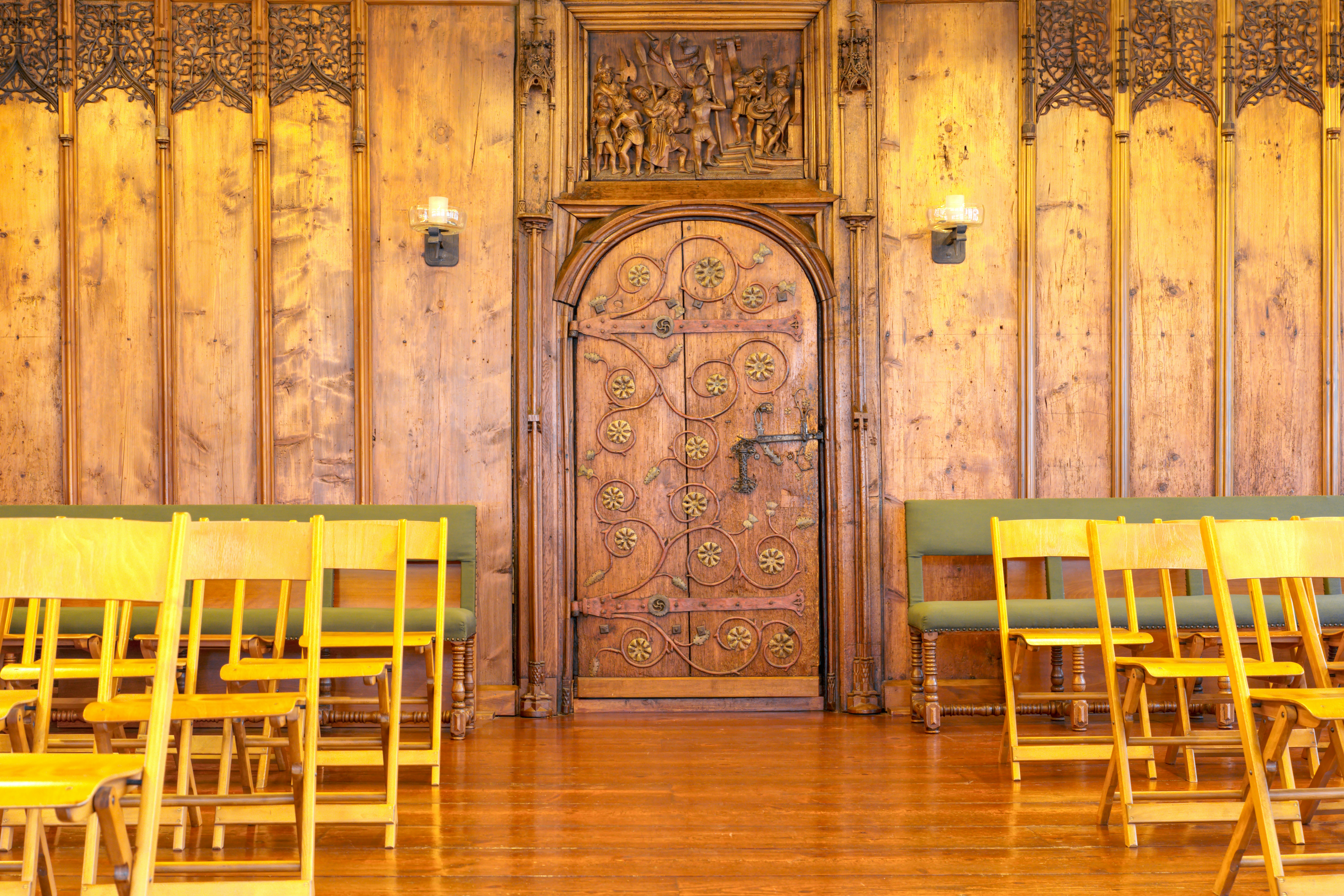 Warmly lit wooden doorway framed by carved panels with yellow chairs along the sides of a polished floor.