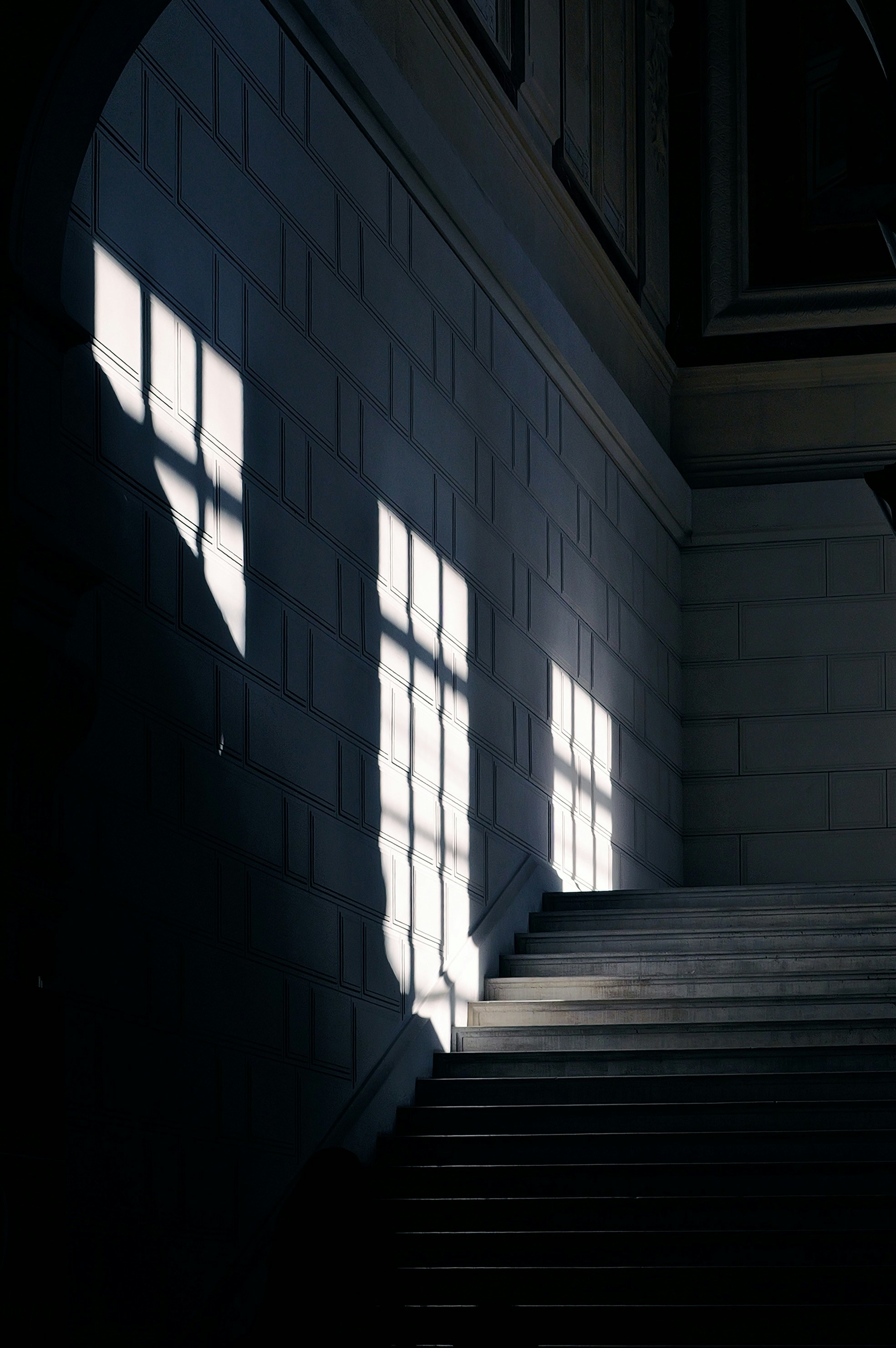 Sunlit window shadows carve geometric patterns along a dim stairwell of a classical interior, highlighting brickwork panels and architectural depth.