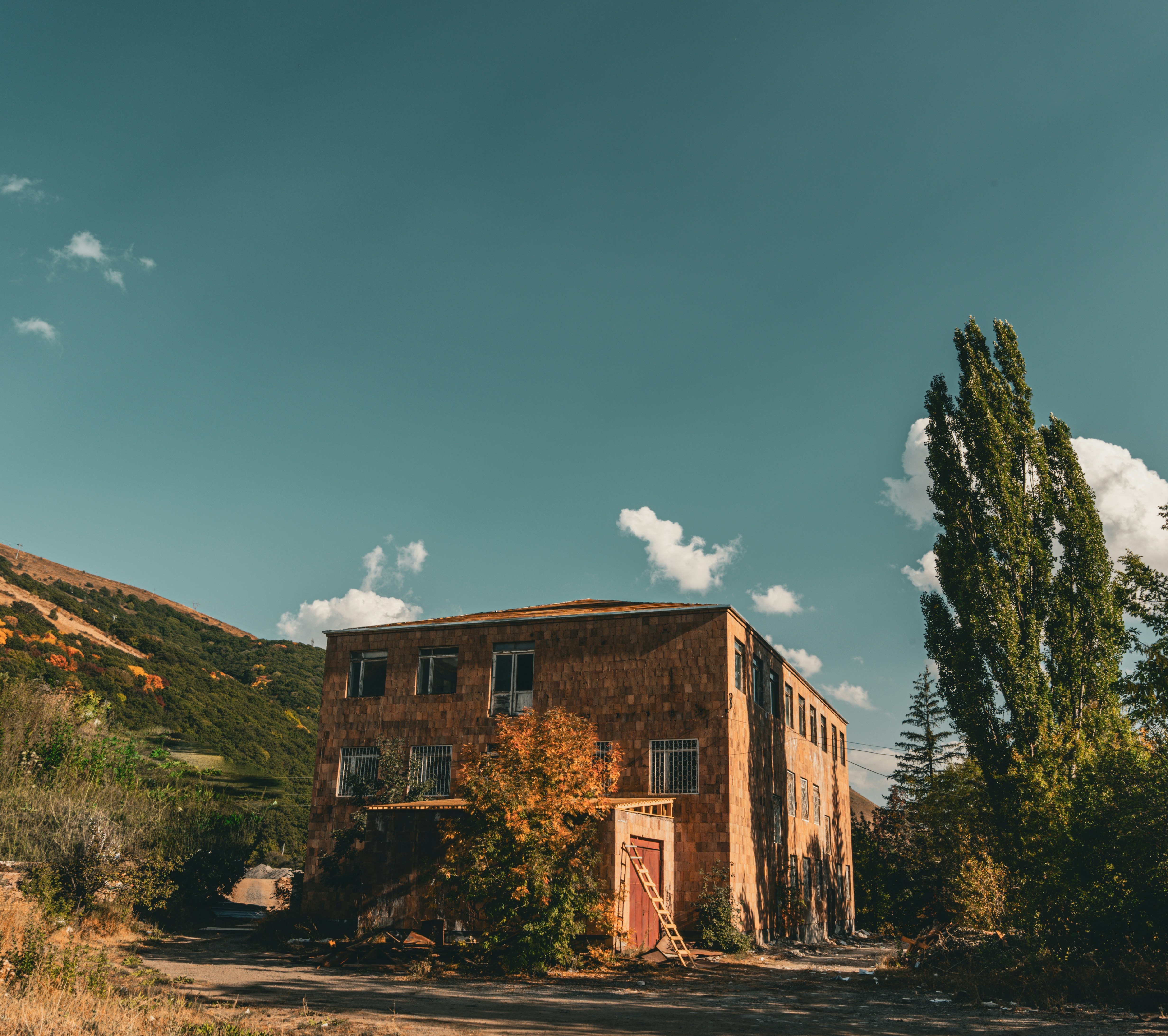 Deserted stone building sits among autumn foliage and dusty paths, anchored by a tall tree on the right and distant hills under a bright sky.