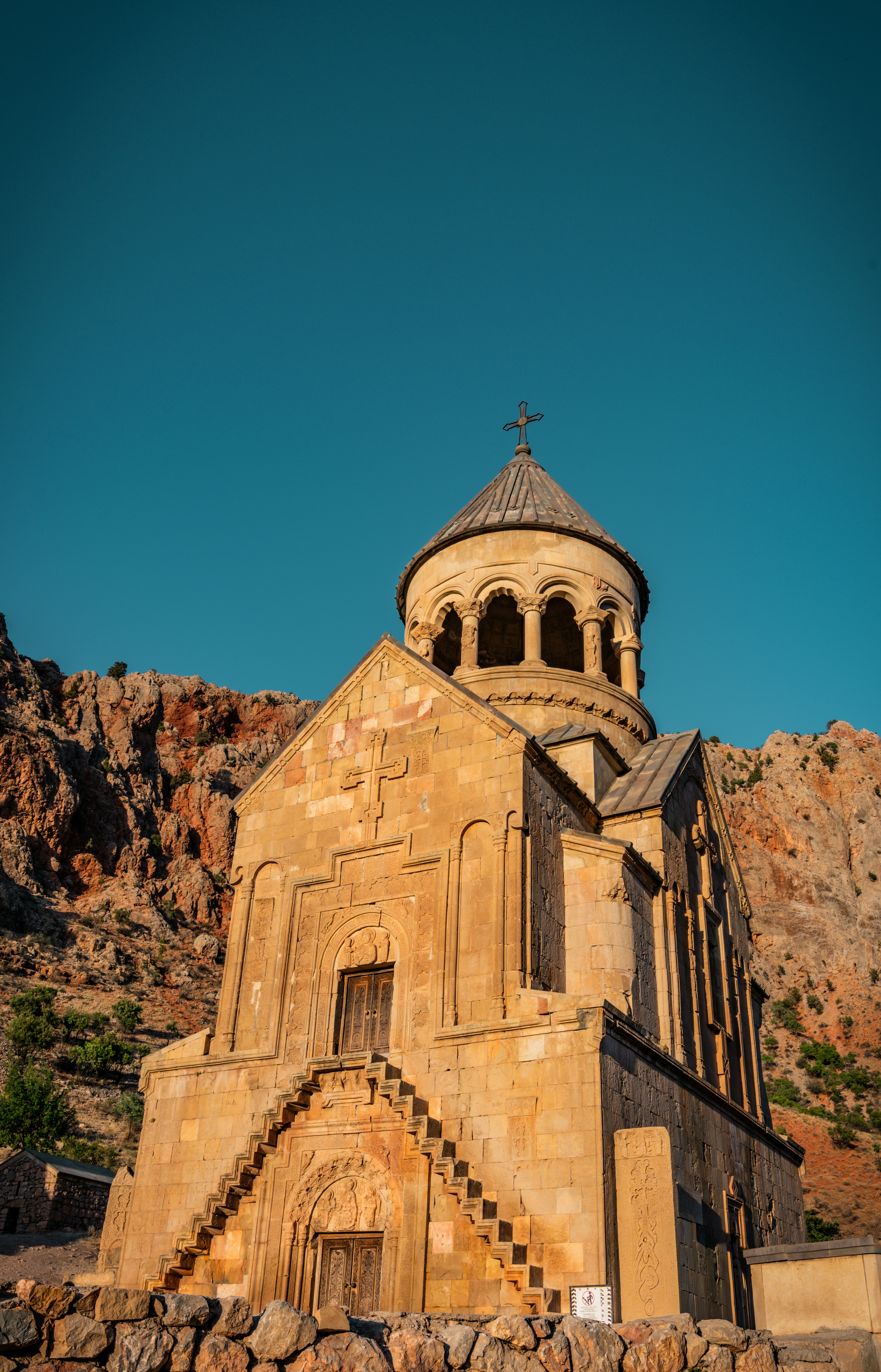 Ancient stone chapel perched against rocky cliff with a domed tower under a clear blue sky. The structure's warm hues contrast with the deep teal backdrop, highlighting architectural details.