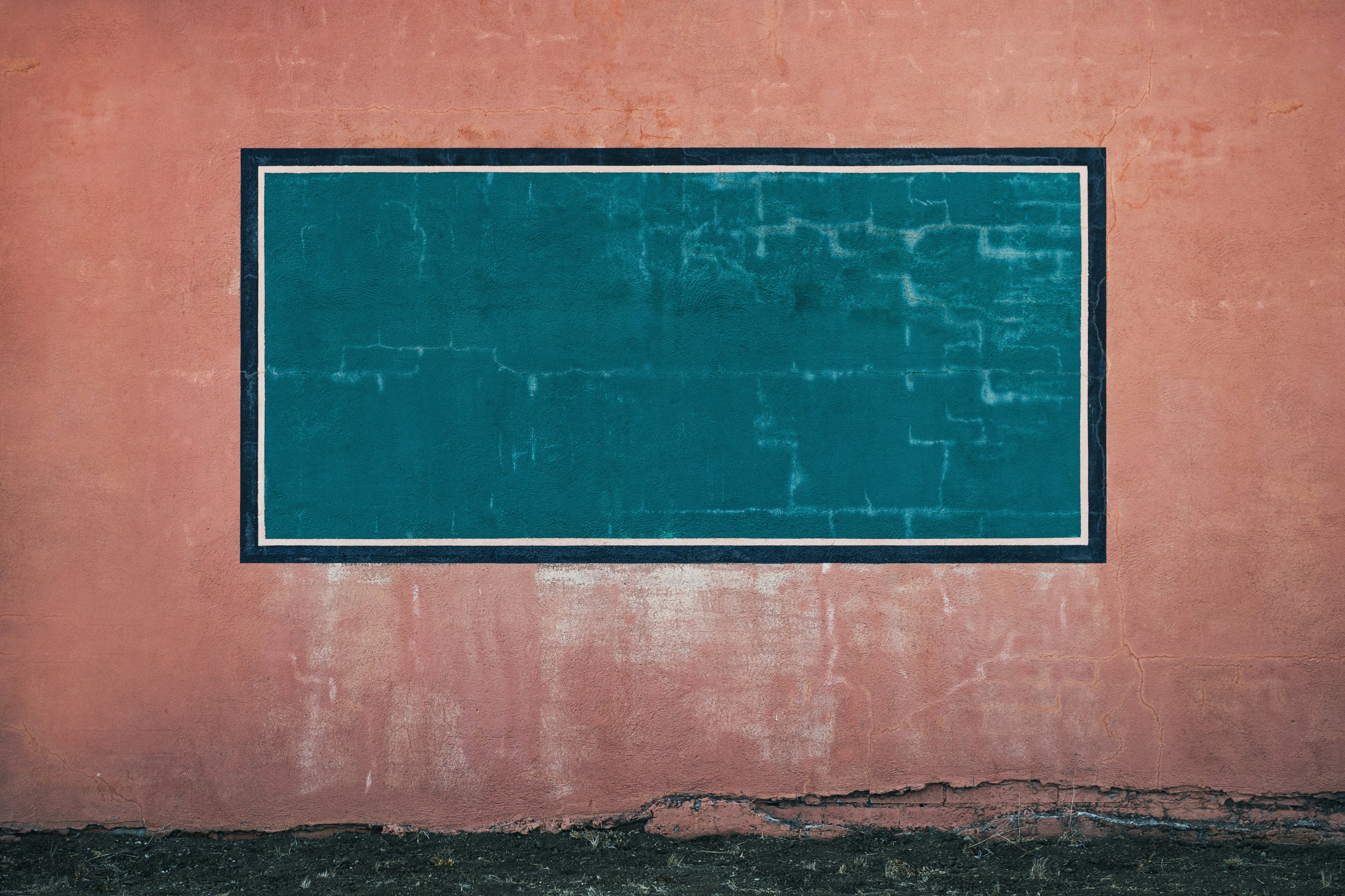 Teal rectangular frame on a sun-washed pink wall, with a dark bottom edge and visible texture; a muted urban surface study featuring a single, centered architectural element.