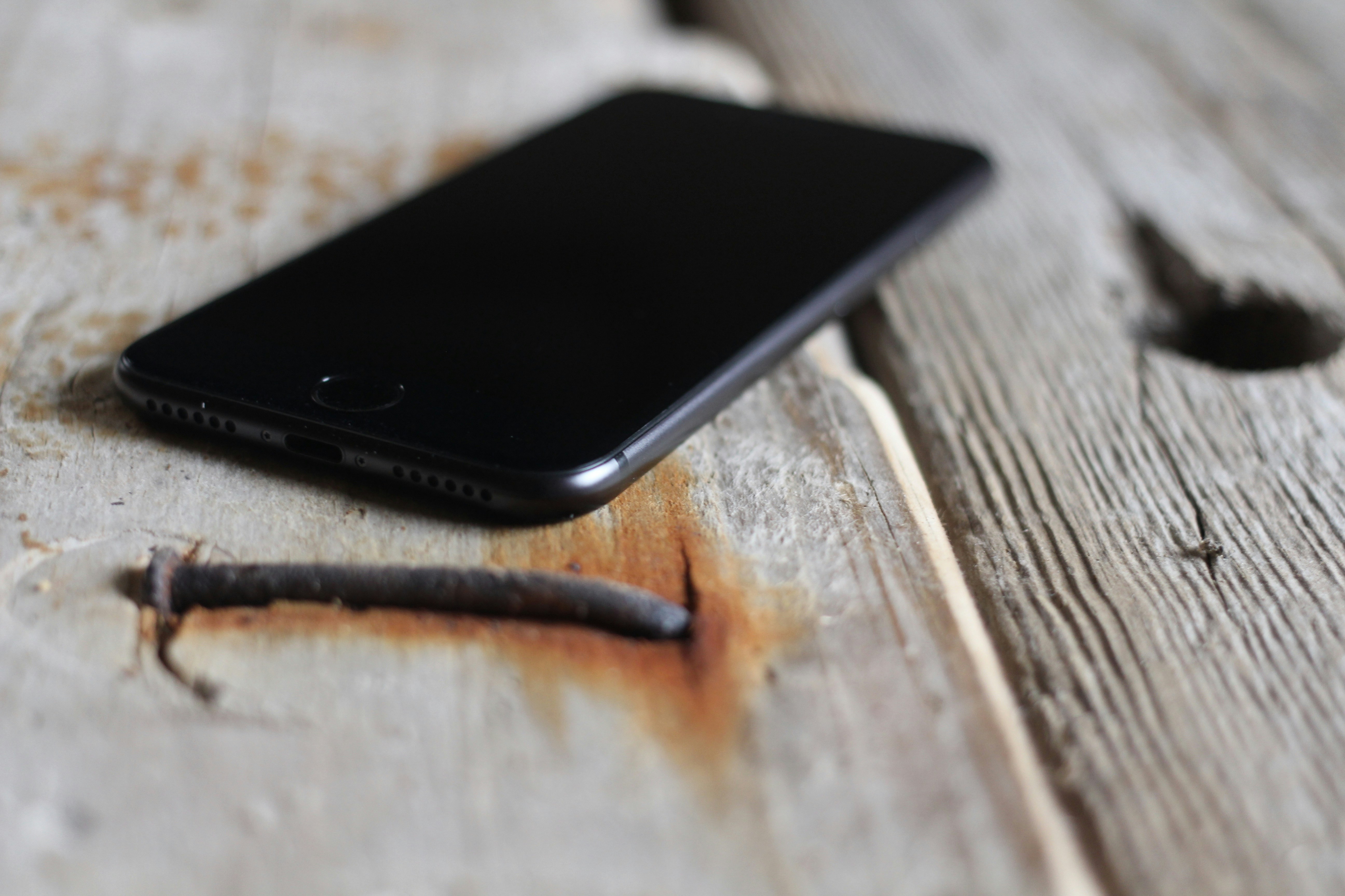 A black smartphone rests by a rusted nail on a wooden surface. The lighting is dim, creating a somber atmosphere.