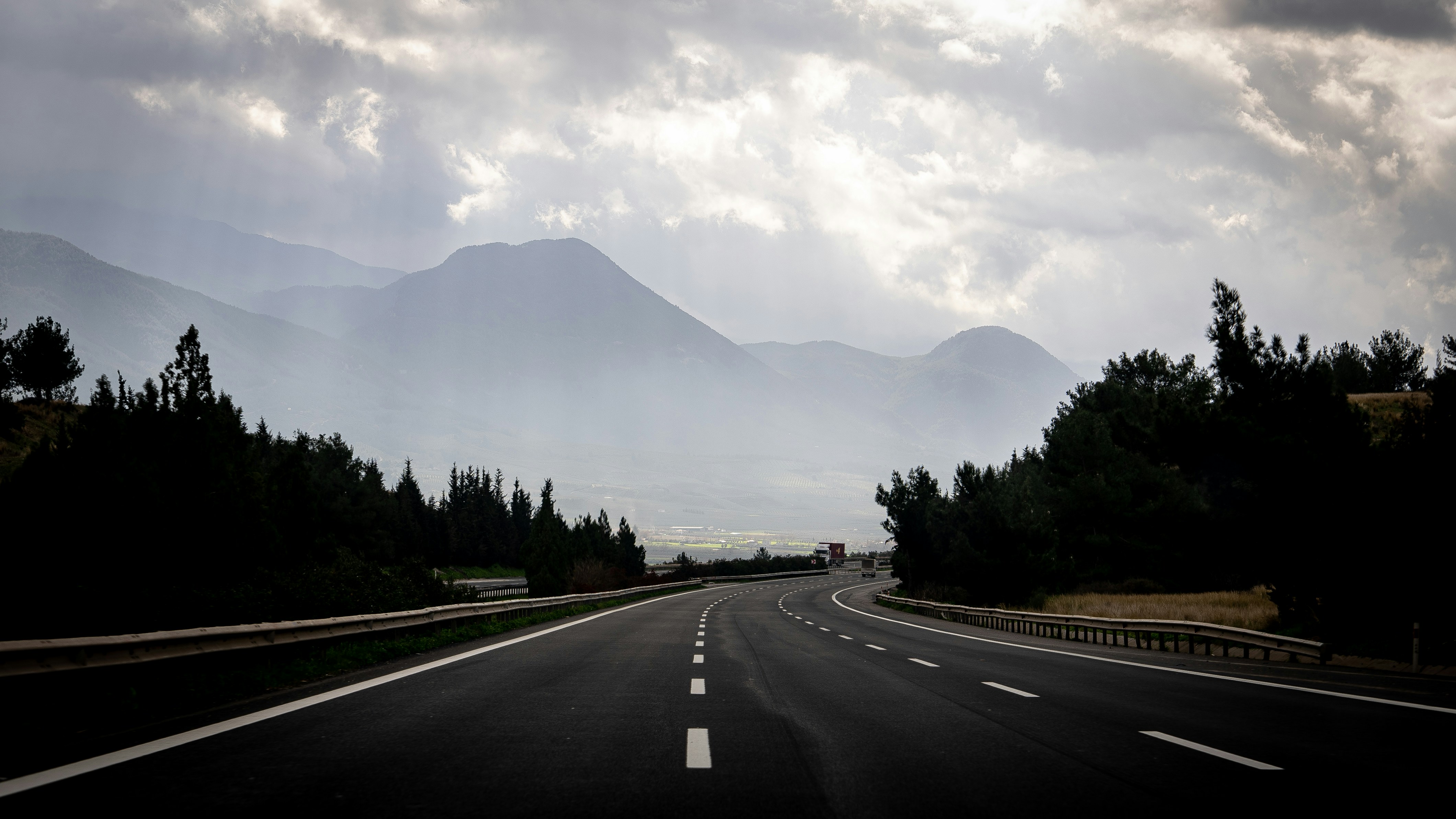 Long, winding highway cuts through a misty valley with silhouettes of conifers and layered mountains in the distance; the scene conveys depth and a sense of journey, with a moody sky above.
