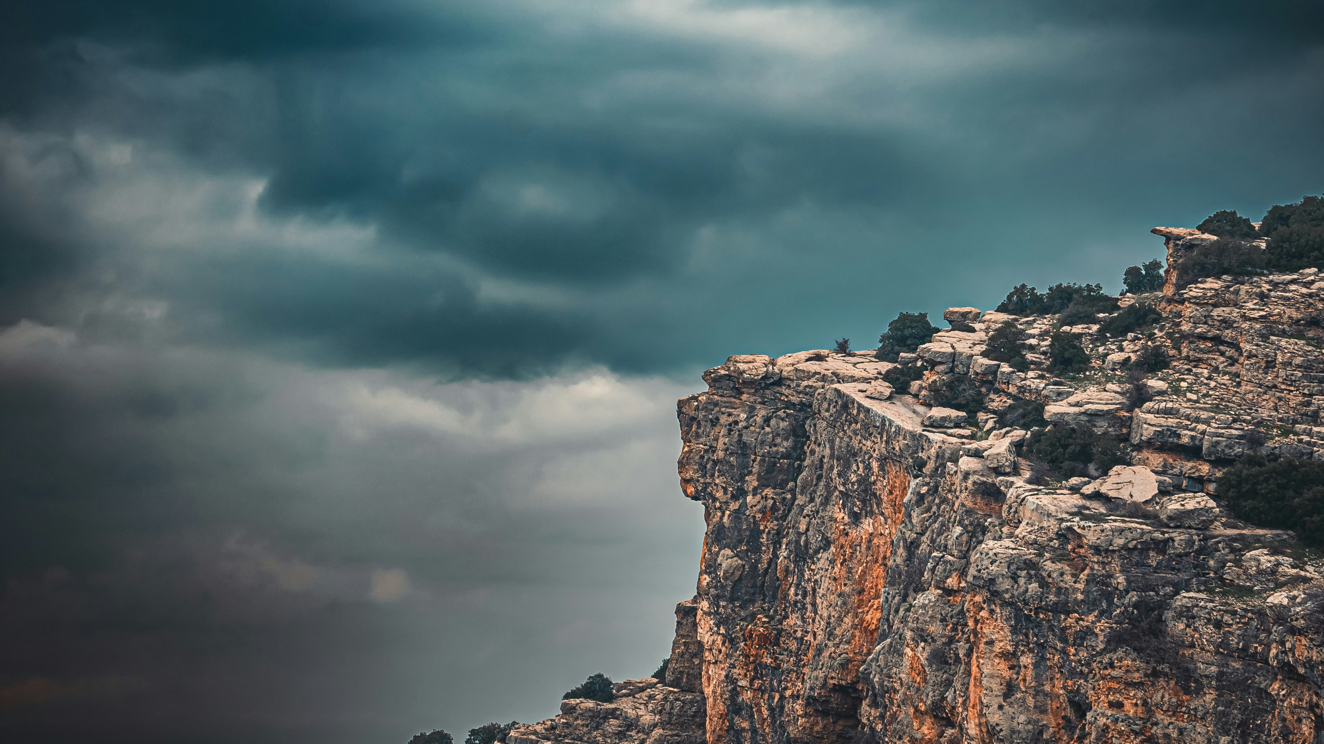 Rugged coastal cliff with stratified rock faces under moody, turbulent clouds; a stark, natural landscape emphasizing texture and scale.
