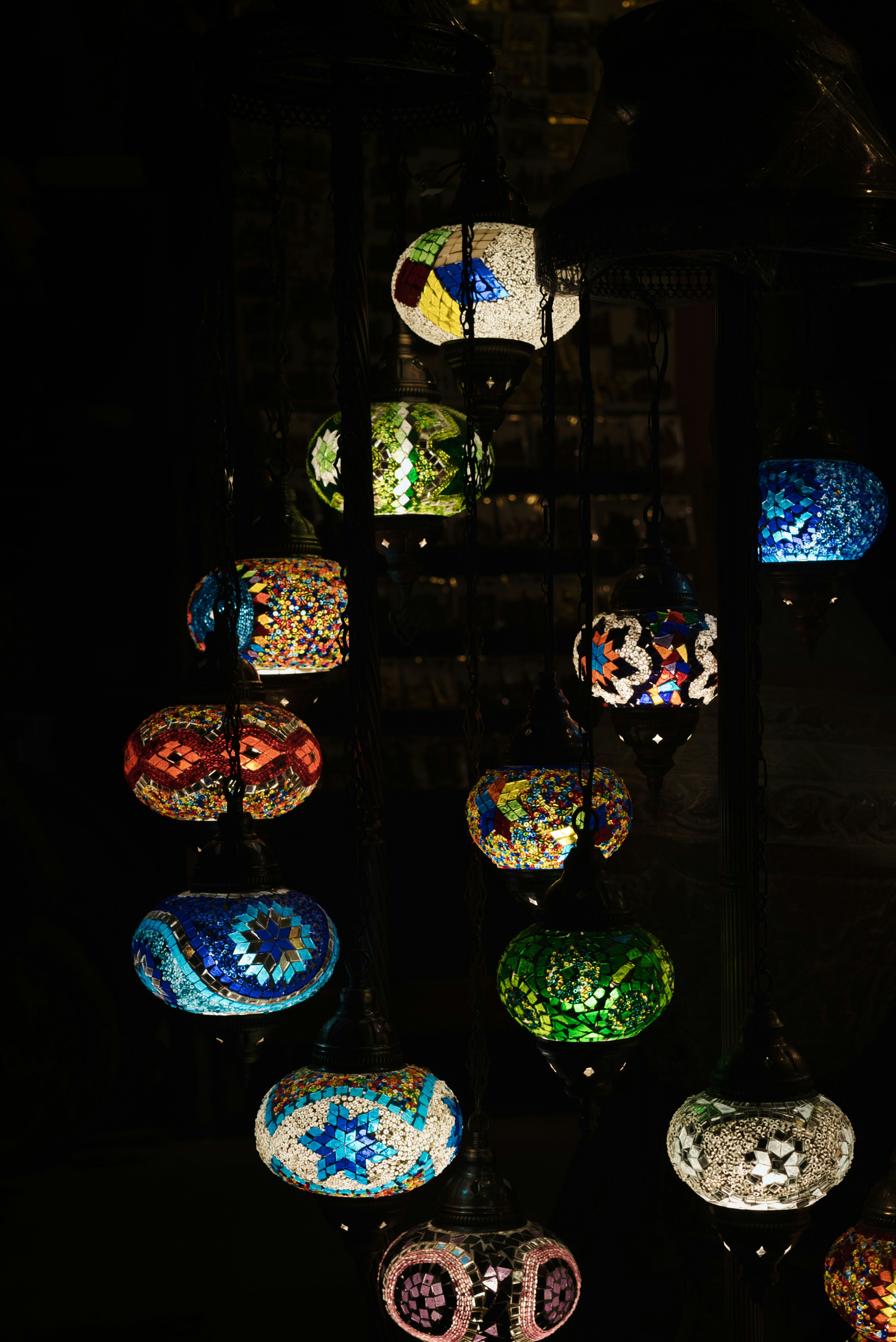 Colorful mosaic lanterns hang in a dim setting, their patterns glowing softly against the dark background. A still life of illuminated glass orbs forming a vertical arrangement.