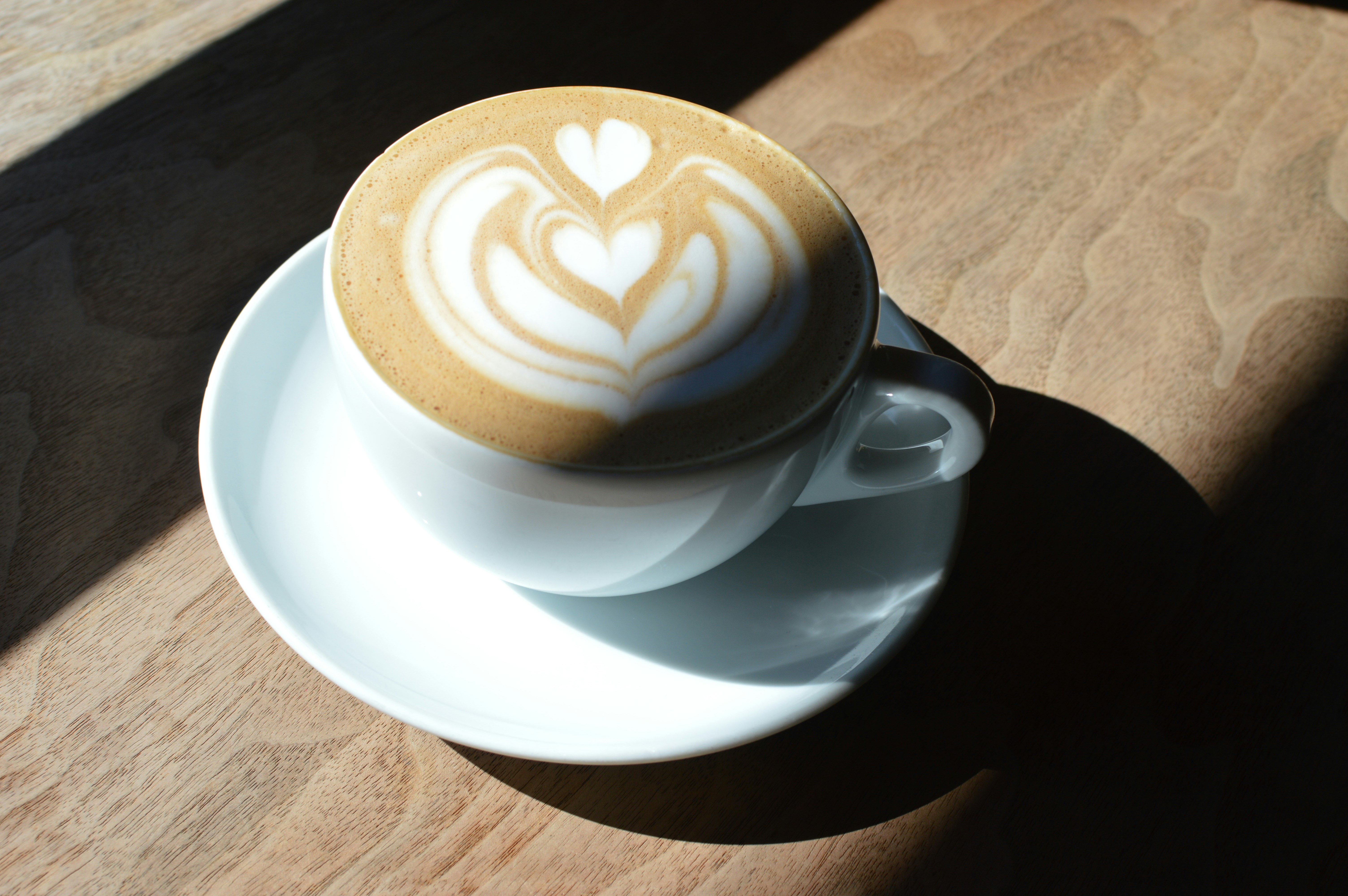 Latte with a heart-shaped foam pattern sits on a white saucer beside a sunlit wooden surface.