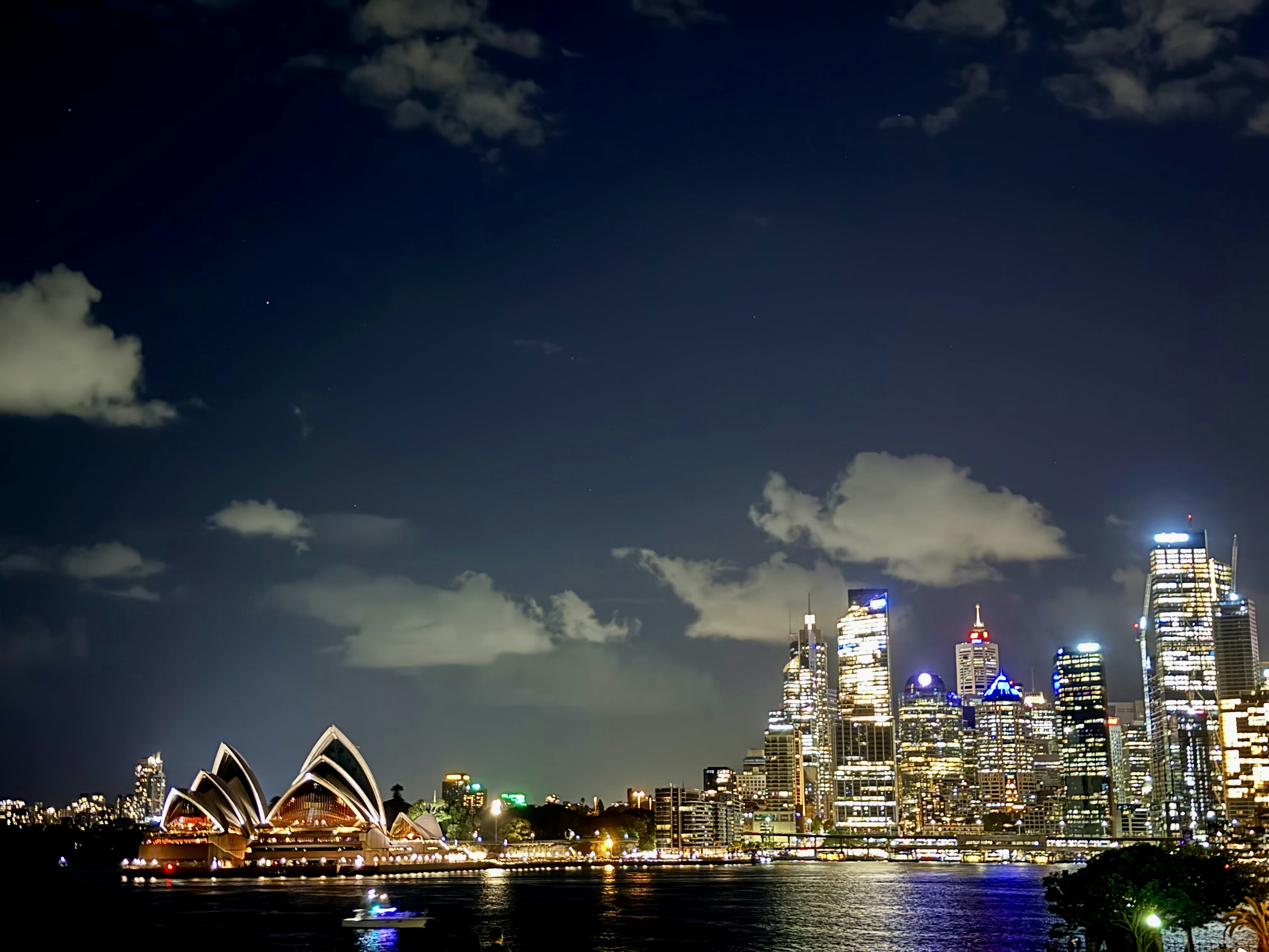Night harbor skyline with the Sydney Opera House illuminated beside a cluster of modern skyscrapers, reflecting on calm water.