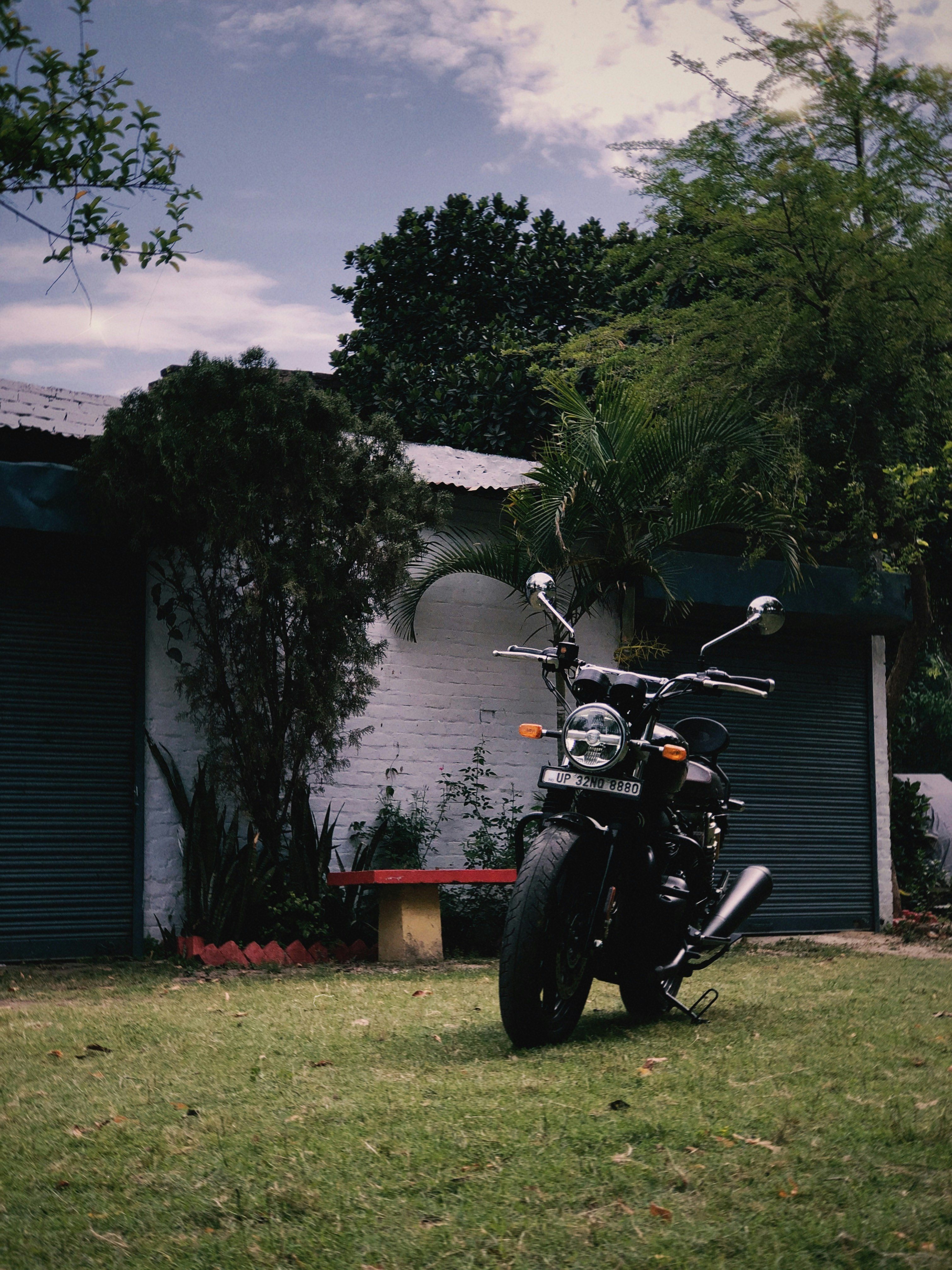 Motorcycle parked on a grassy yard with a brick wall and closed shutters in the background, bathed in natural daylight. The scene emphasizes the bike as the focal point of a quiet outdoor space.