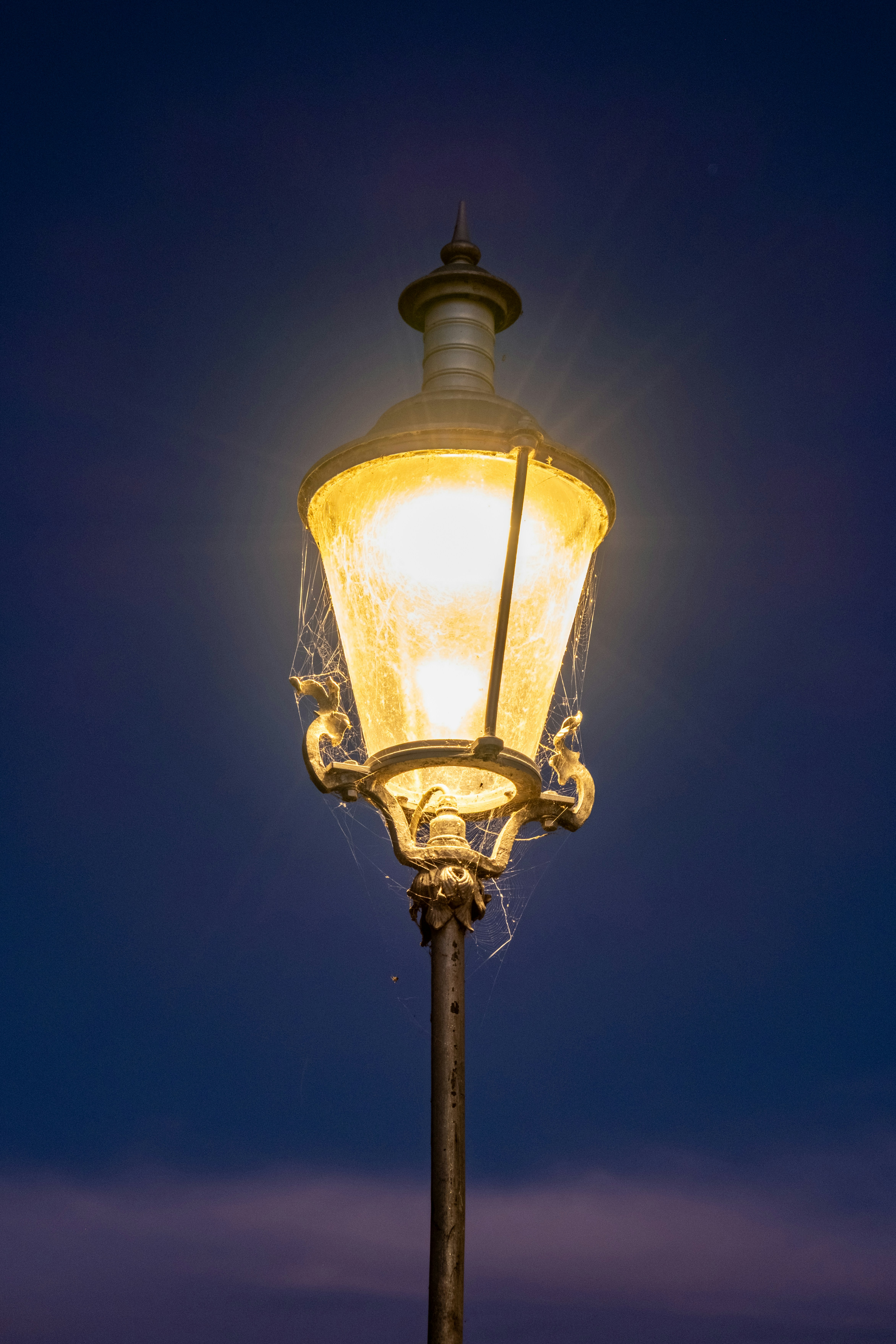 A glowing vintage street lamp against a dark evening sky with spider webs visible around the lantern, emphasizing its warm light and weathered post.