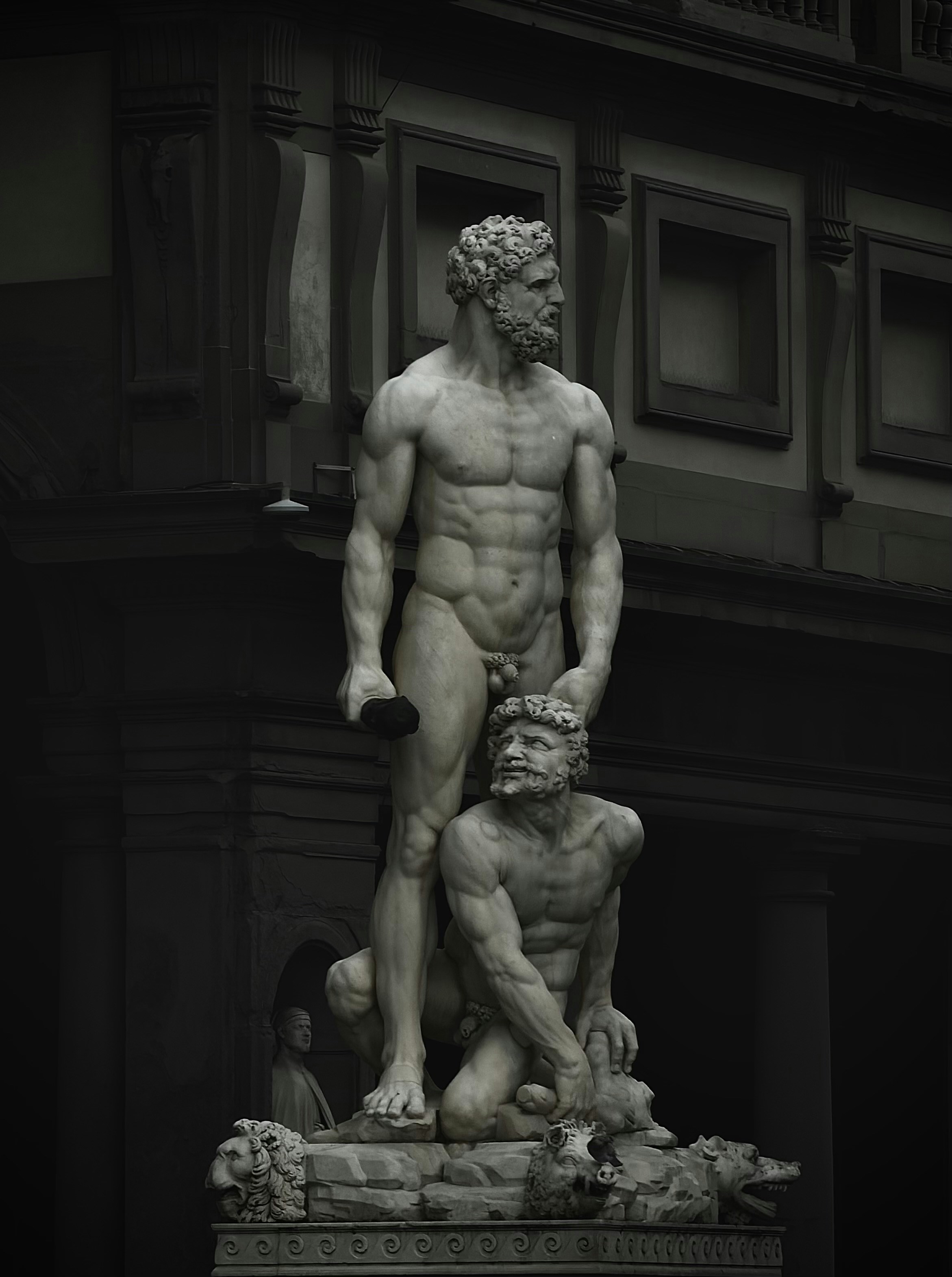Two classical statues stand atop a carved pedestal against a dim architectural backdrop, with the taller figure gripping a hammer while the smaller figure kneels beside him; the scene emphasizes muscular form and sculptural detail.