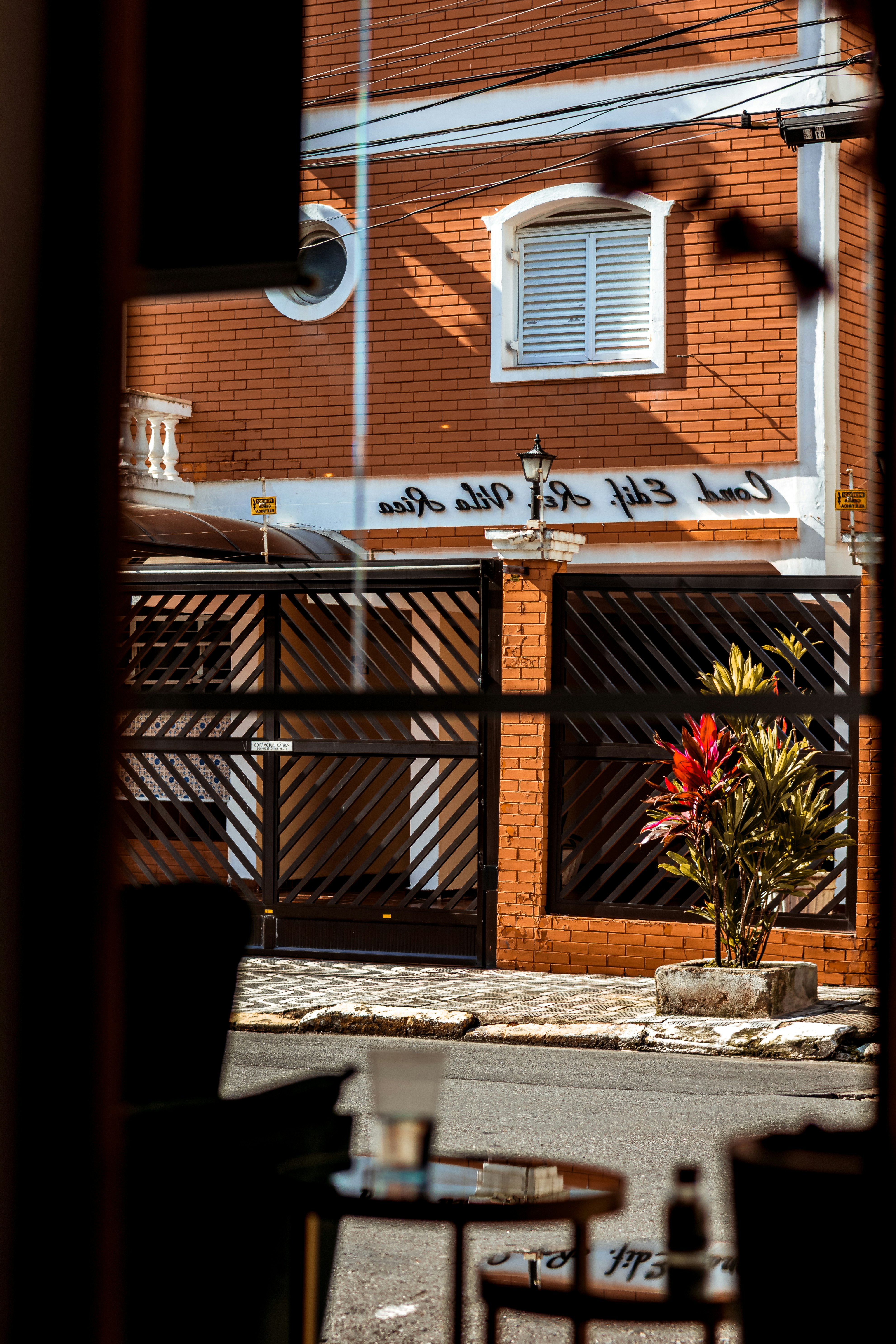 Sunlit brick building with arched window and shutters, viewed through a dark foreground frame; a patterned gate and a potted plant add depth to the street scene.