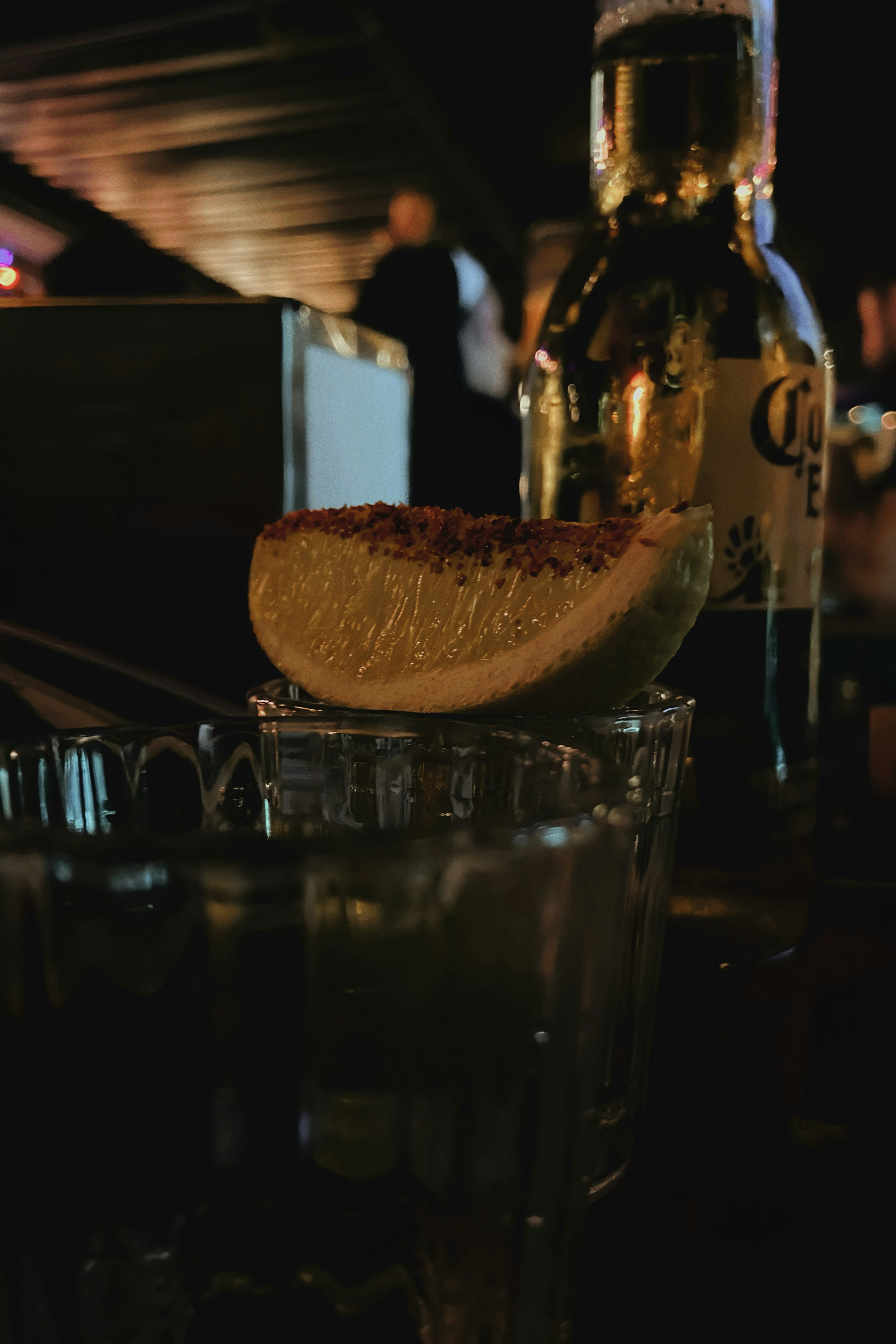 Lime wedge perched on a row of shot glasses with a beer bottle and dark bar backdrop; moody lighting emphasizes texture and glow of citrus and glass.