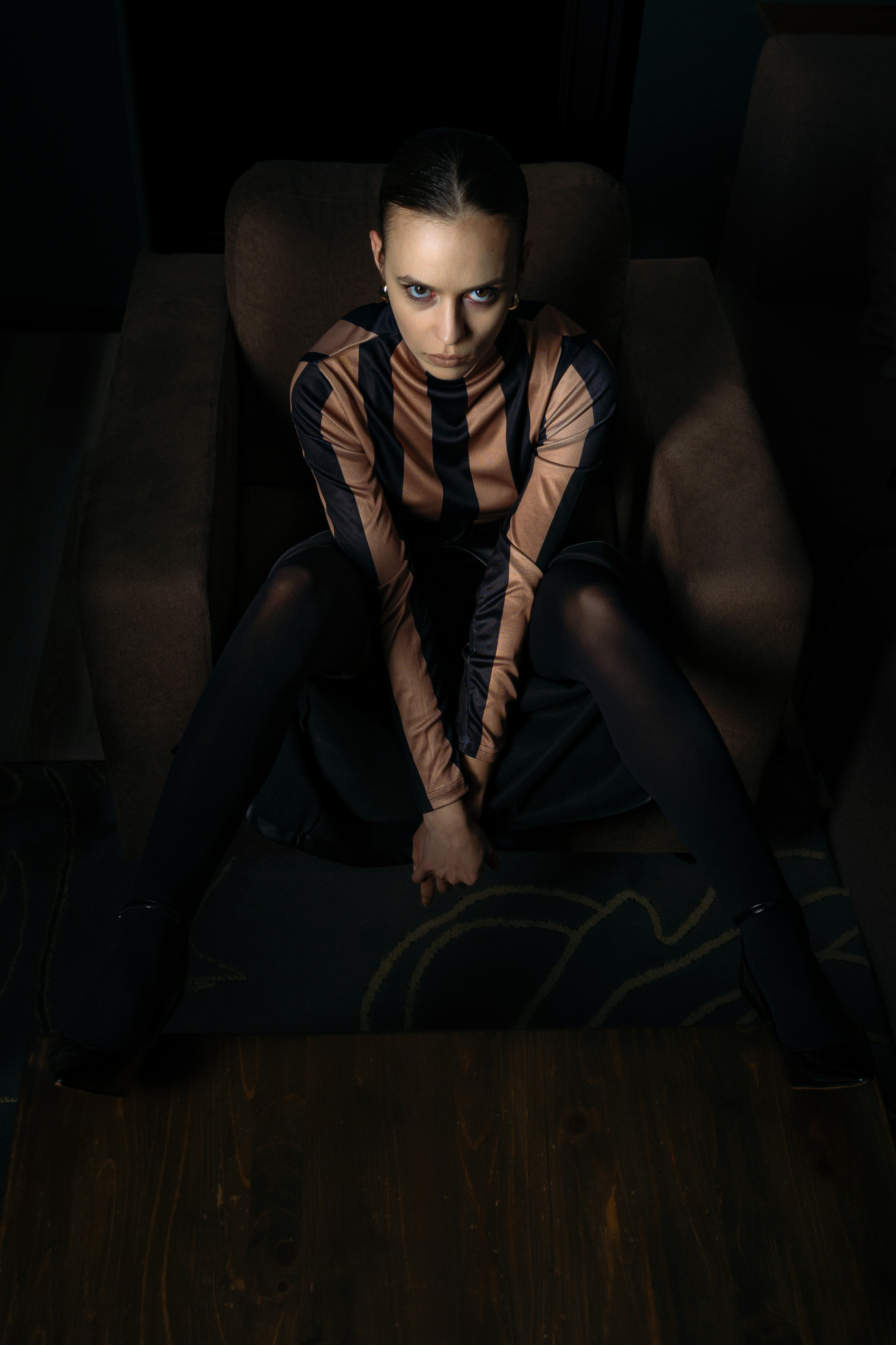 A woman sits in a dark parlor wearing a striped dress, knees drawn up as shadows frame her focused gaze. The scene emphasizes mood and form as a fashion portrait.
