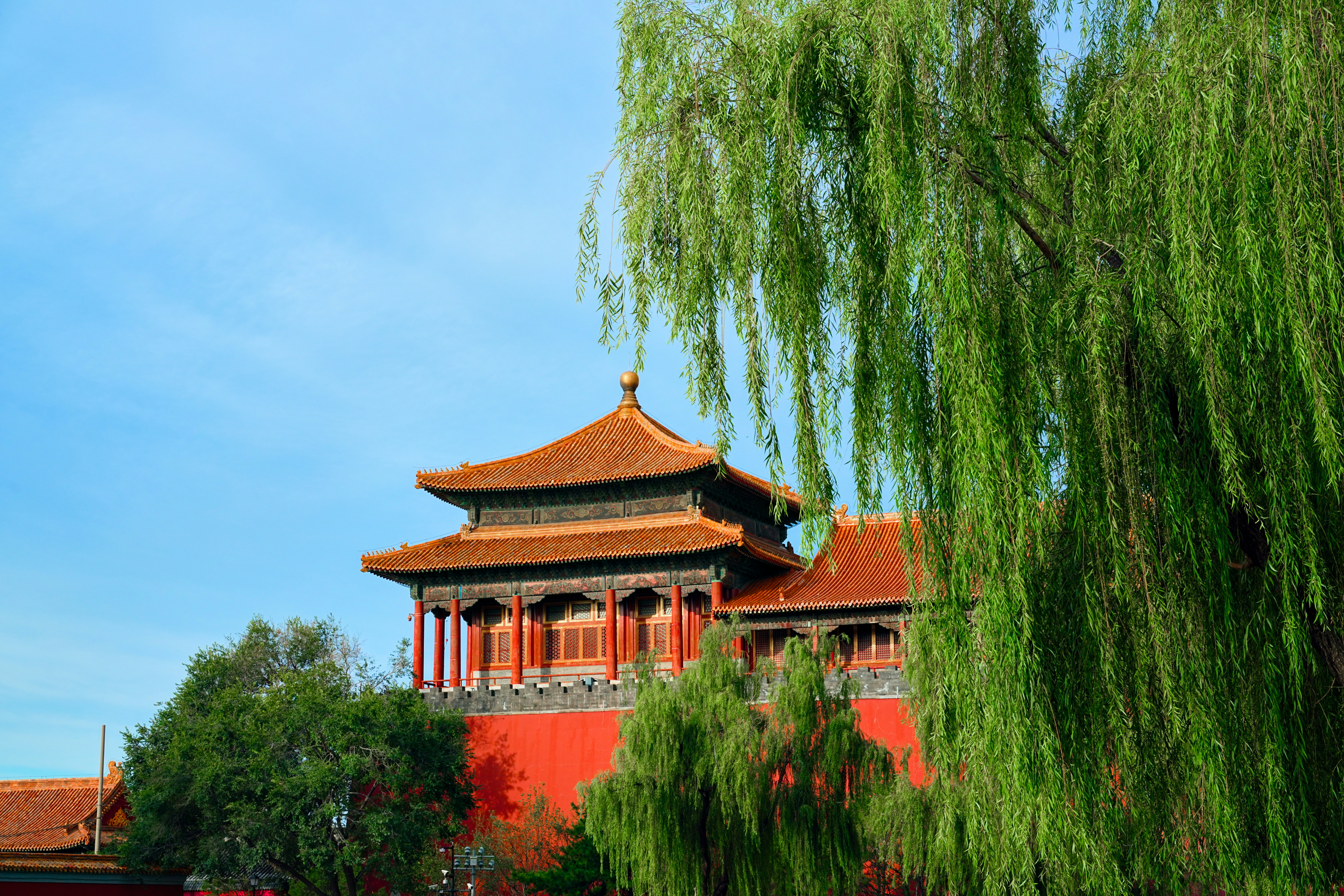 Bright red traditional building framed by overhanging willow branches against a clear blue sky. A calm landscape with architectural detail and lush greenery.