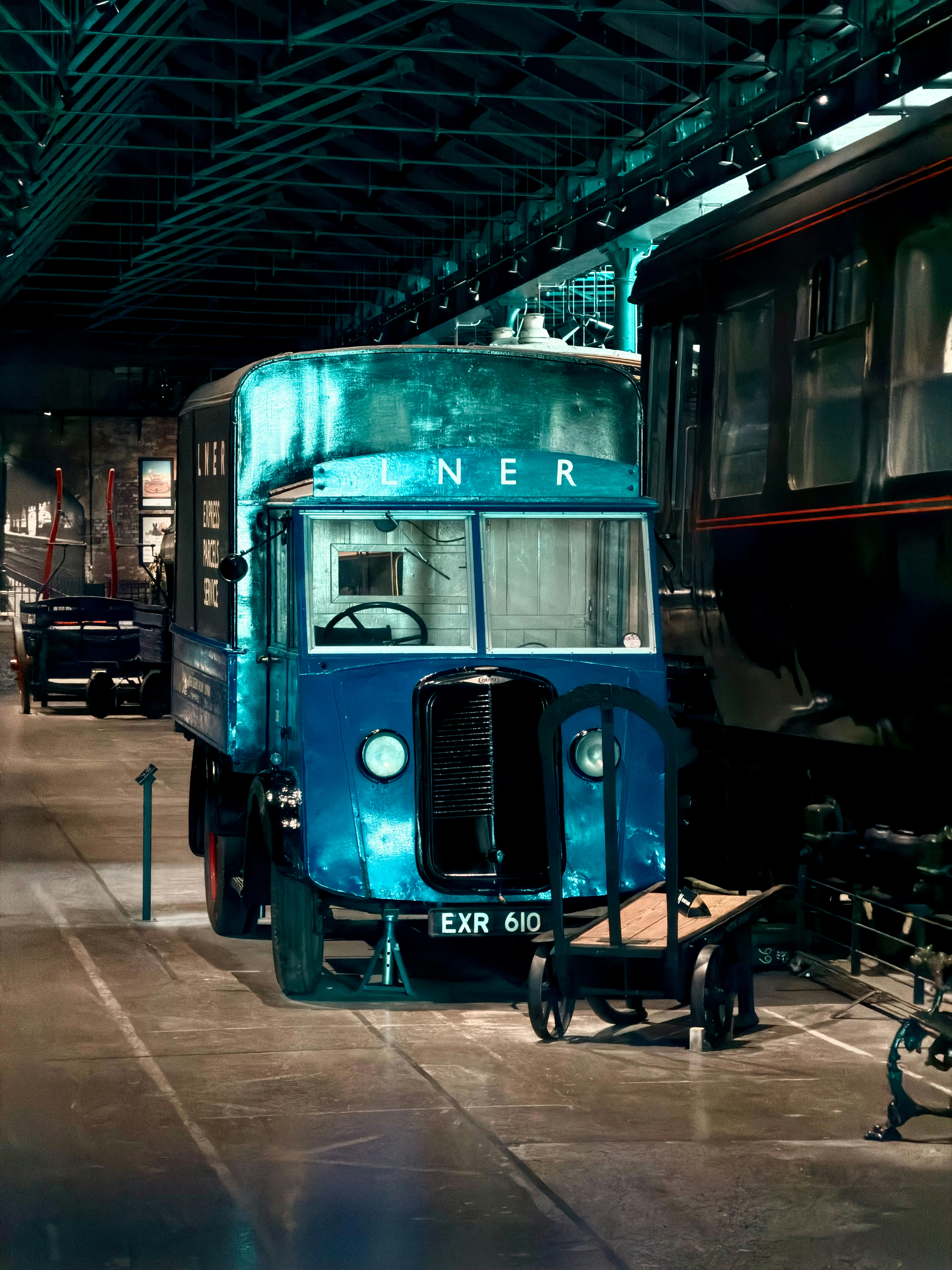 Blue vintage bus/locomotive on display under moody museum lighting, surrounded by trains and equipment; the scene emphasizes industrial heritage inside the gallery-like space.