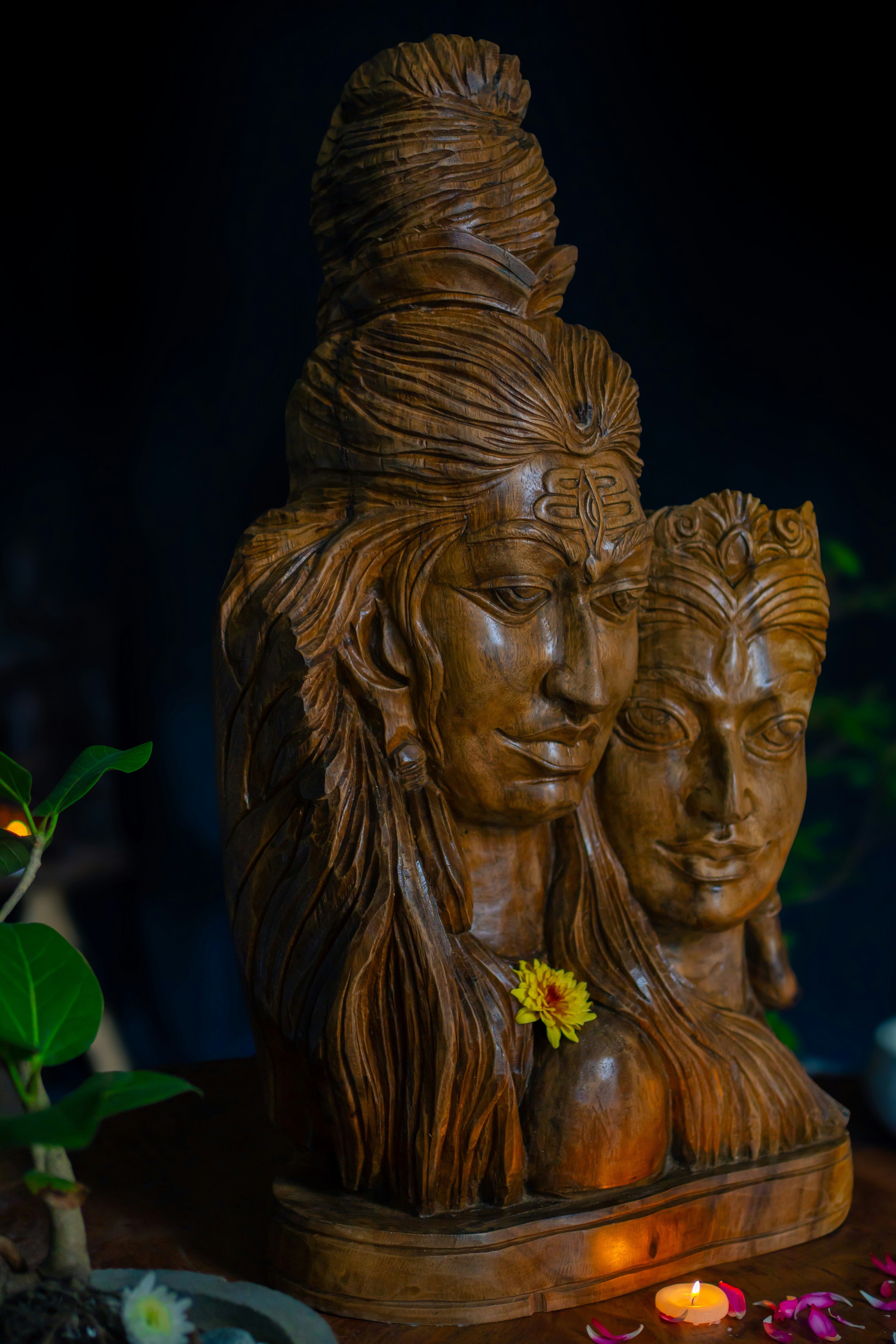Carved wooden statues of two faces sit in soft candlelight with a small flower on the base, accompanied by green leaves in the foreground. A calm, contemplative scene highlighting craftsmanship and tranquil ambience.