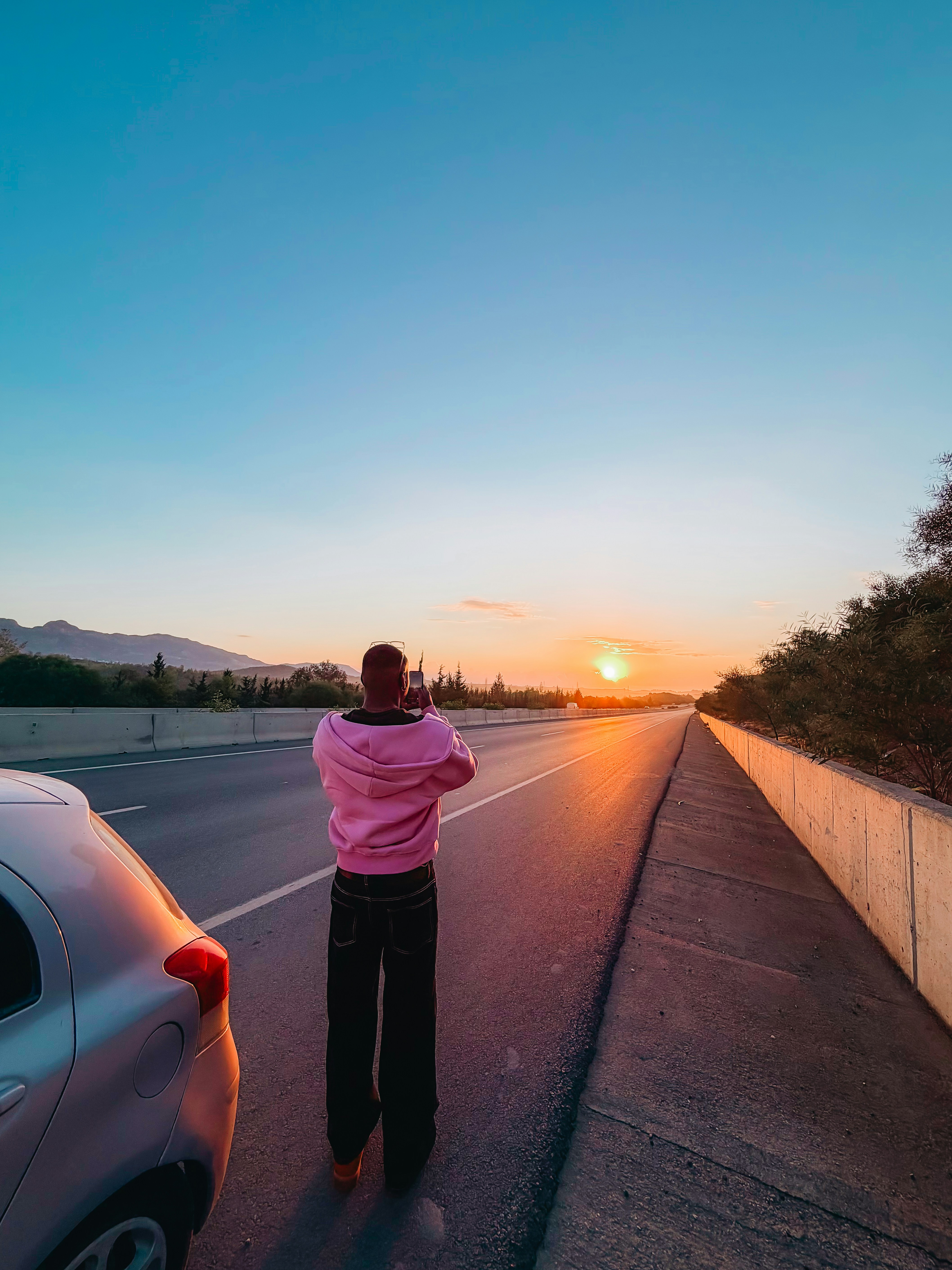 A person in a pink hoodie stands on a roadway shoulder, facing a low sun as the highway stretches ahead with a car parked on the left and trees lining the right. This is a candid street_photo capturing a sunset moment.