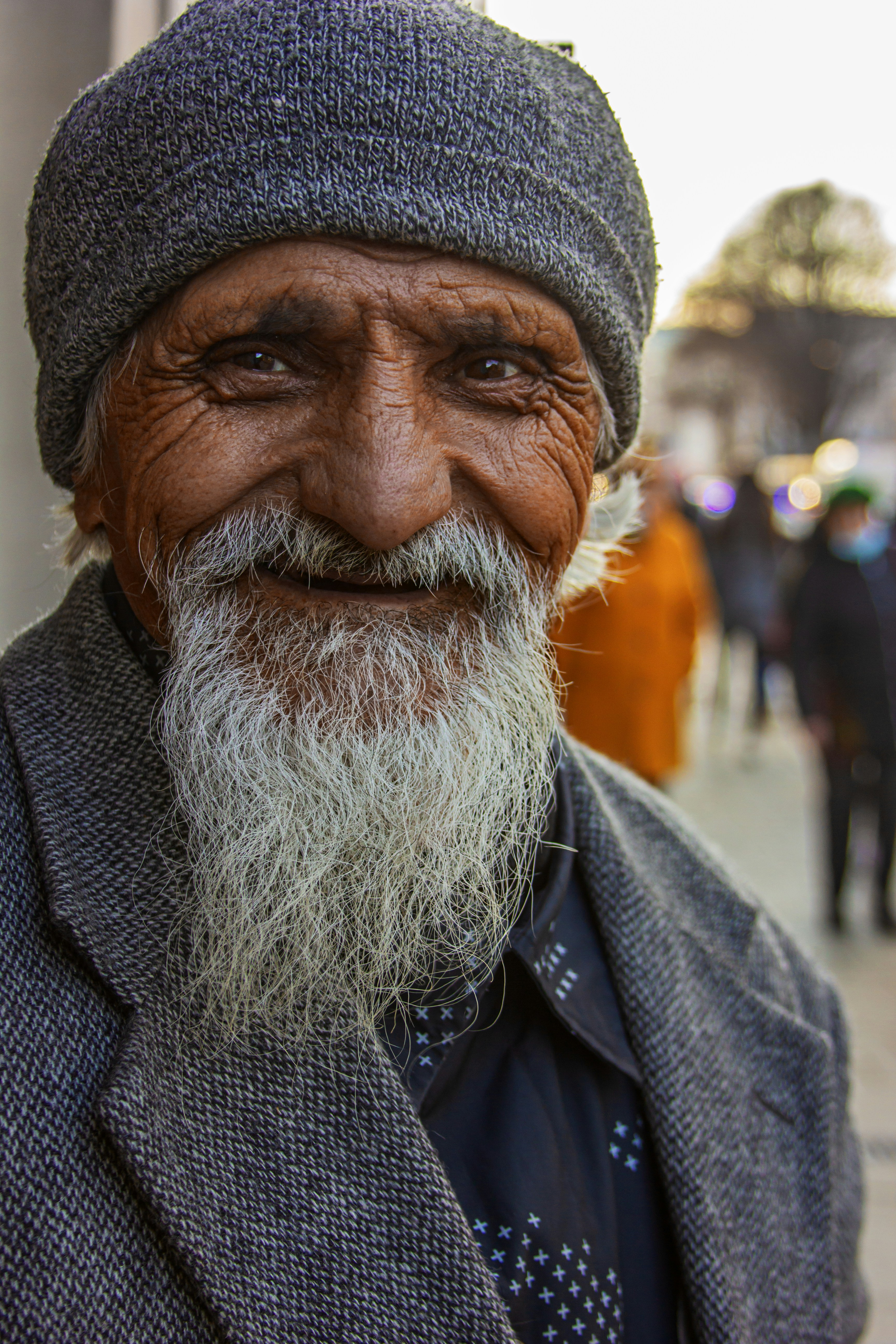 Smiling elderly man with a gray beard and knit cap in an outdoor street scene; focus on facial expression and texture, with blurred background indicating a busy urban environment.