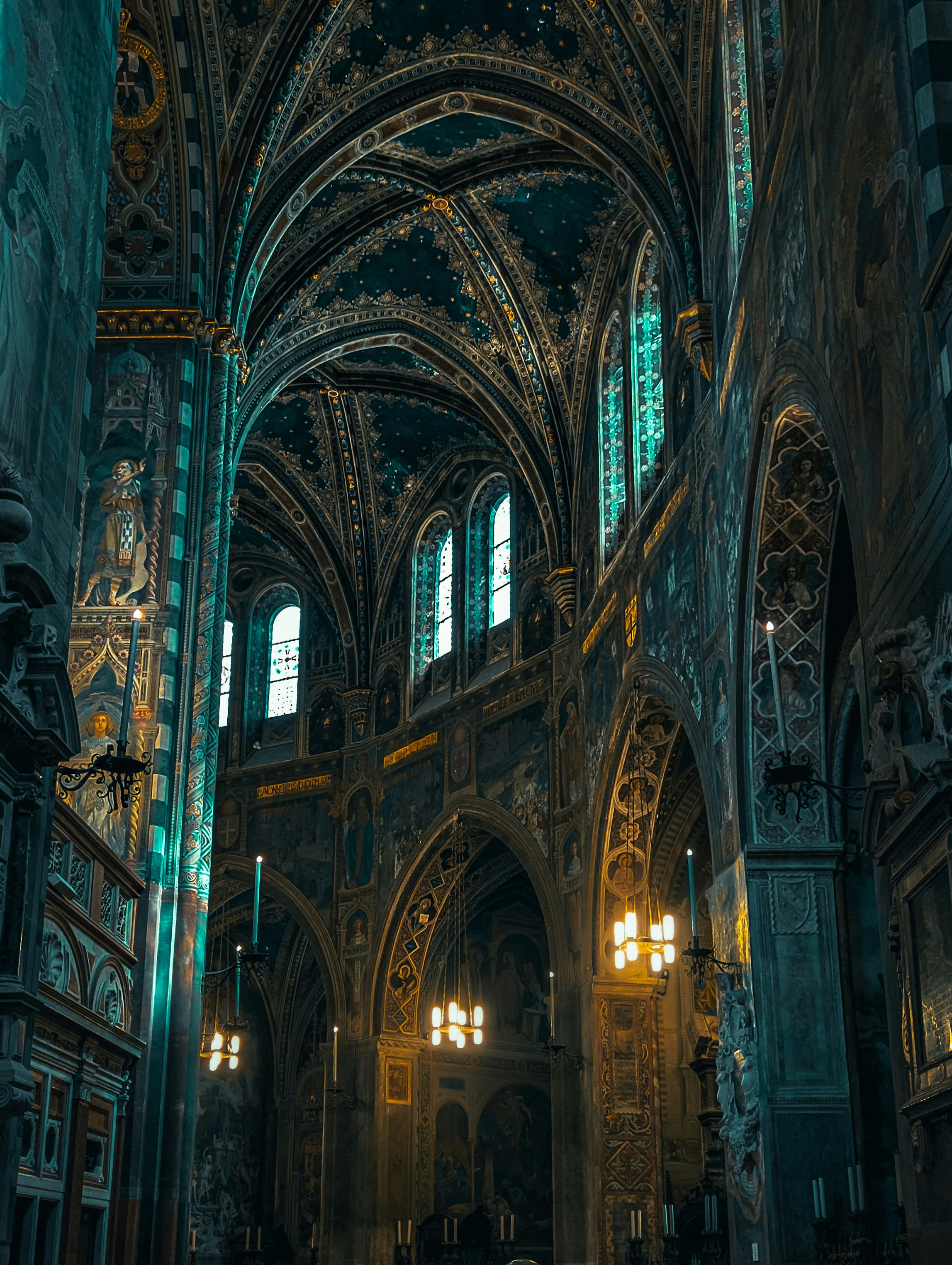 Ornate cathedral interior bathed in teal light, featuring soaring ribbed vaults, stained windows, and glowing chandeliers that illuminate intricate stone carvings.