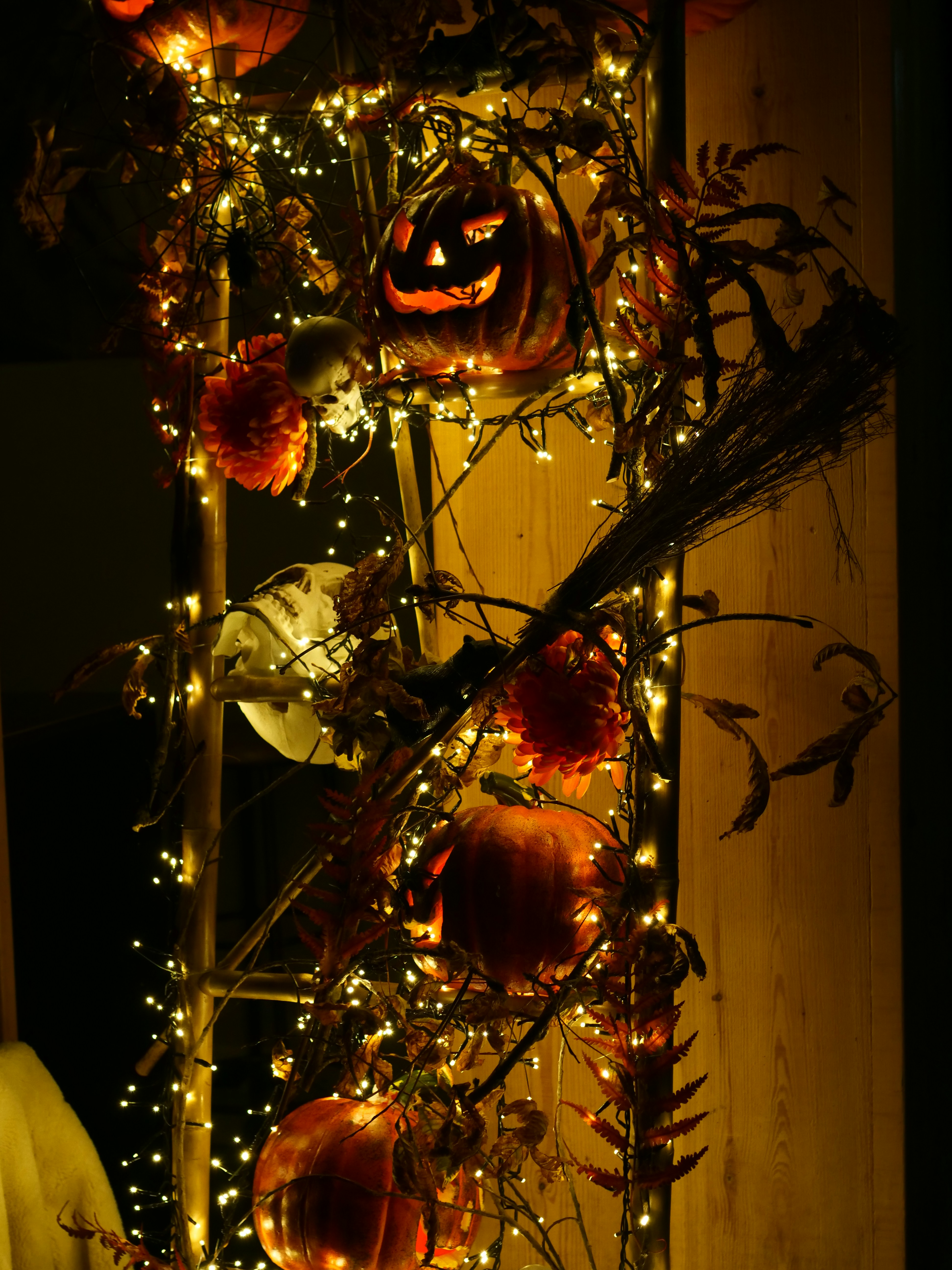 A cozy Halloween centerpiece with carved pumpkins, fairy lights, and autumn foliage arranged along a vertical frame; warm amber tones and soft bokeh highlight the festive display.