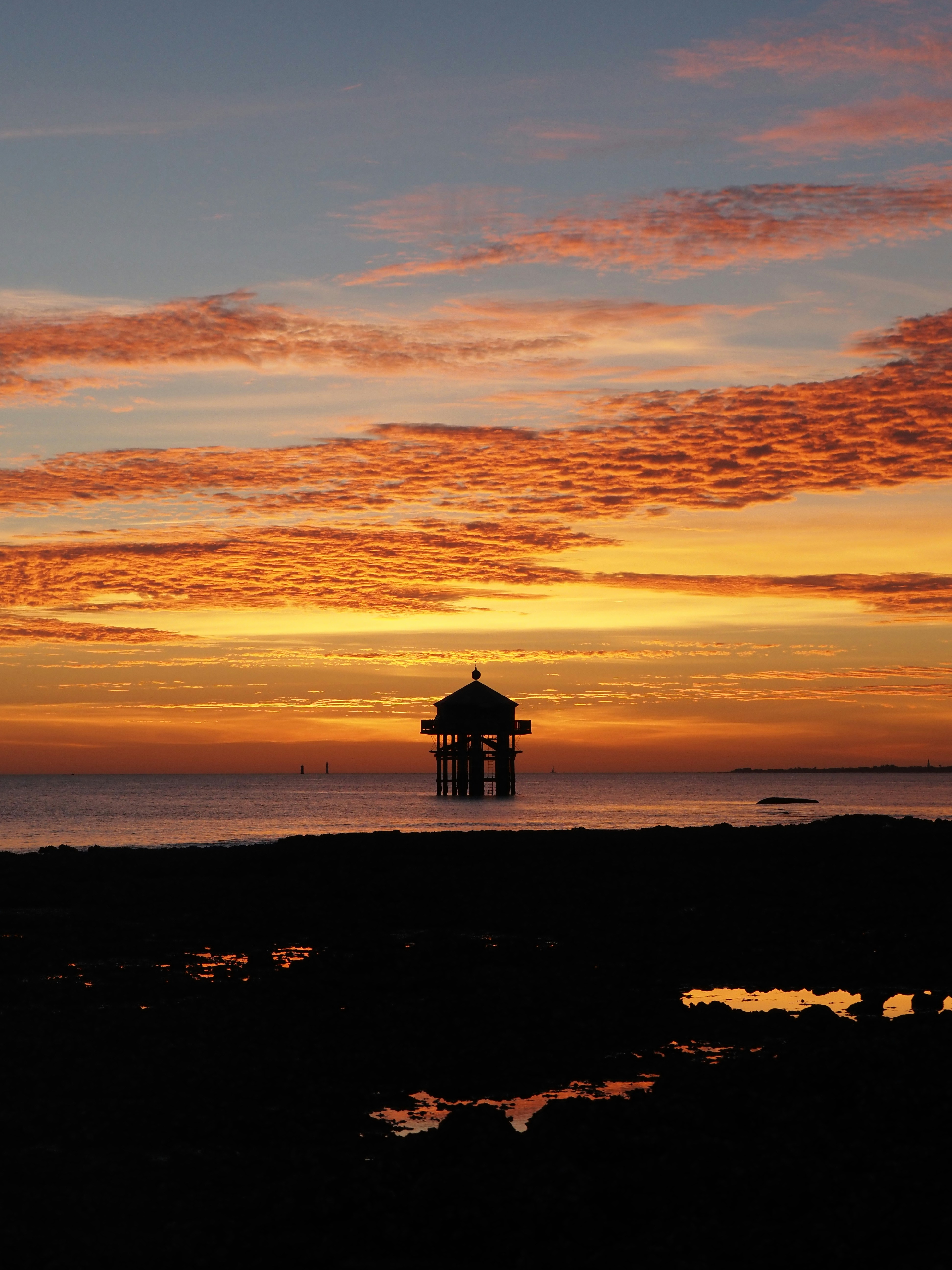 Silhouette of a small pier standing above calm waters at sunset, with vibrant orange and pink clouds filling the sky.