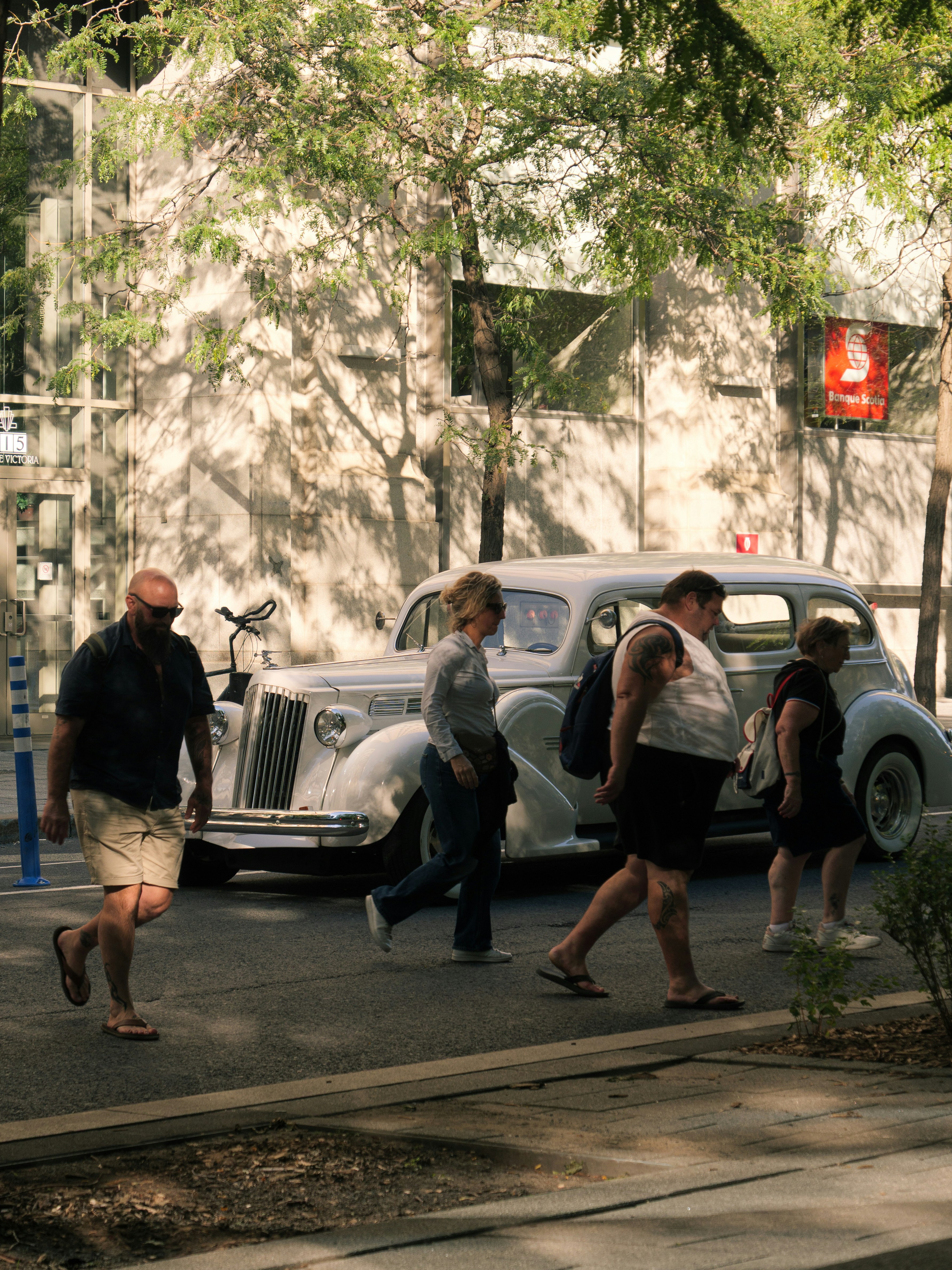 Sun-dappled street scene with a vintage white car as the focal point, and pedestrians passing by in shaded light; a calm urban moment captured on a sunny day.