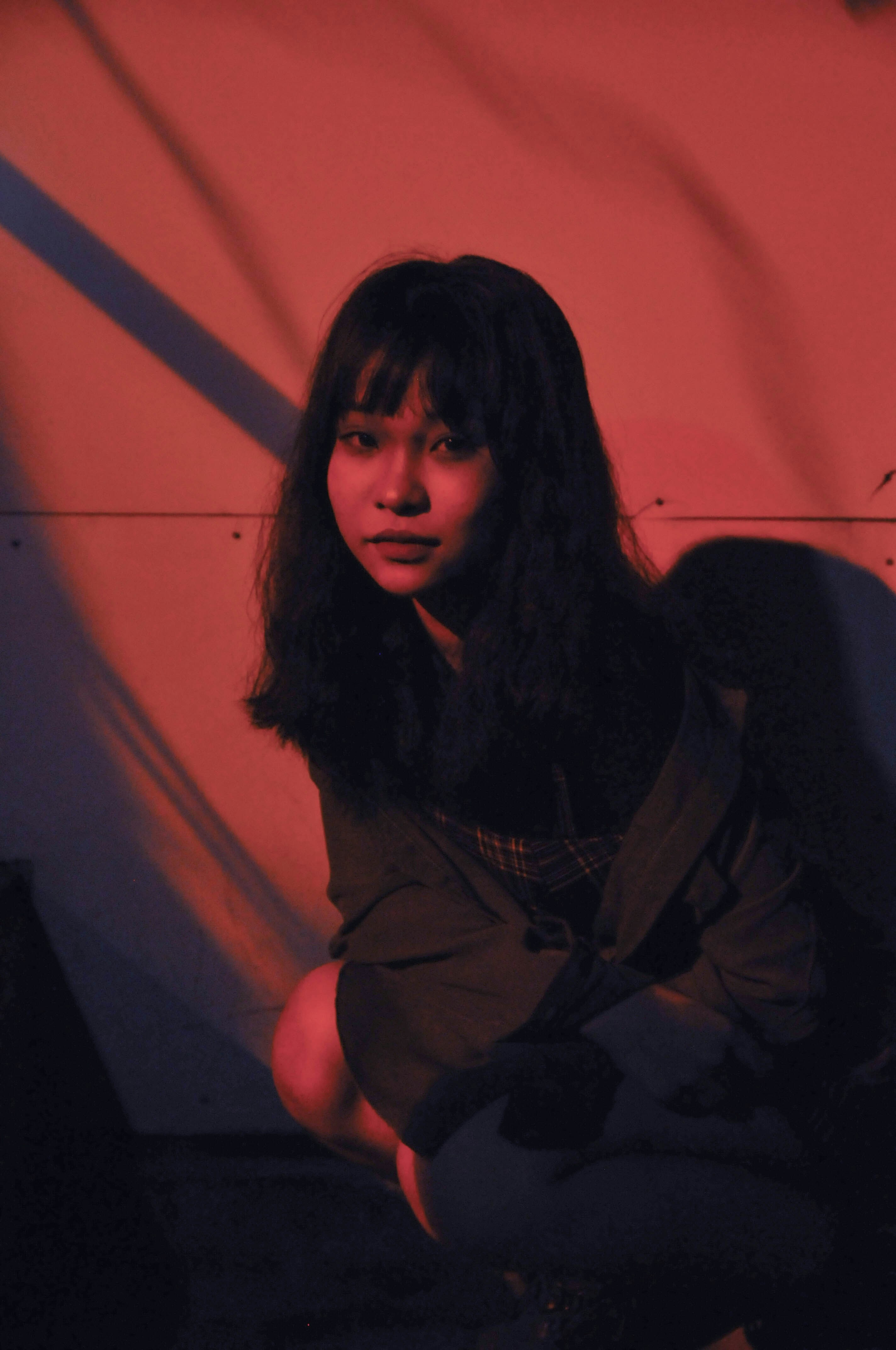 Portrait of a young woman crouching in a dim, red-lit space with soft shadows and moody ambience.