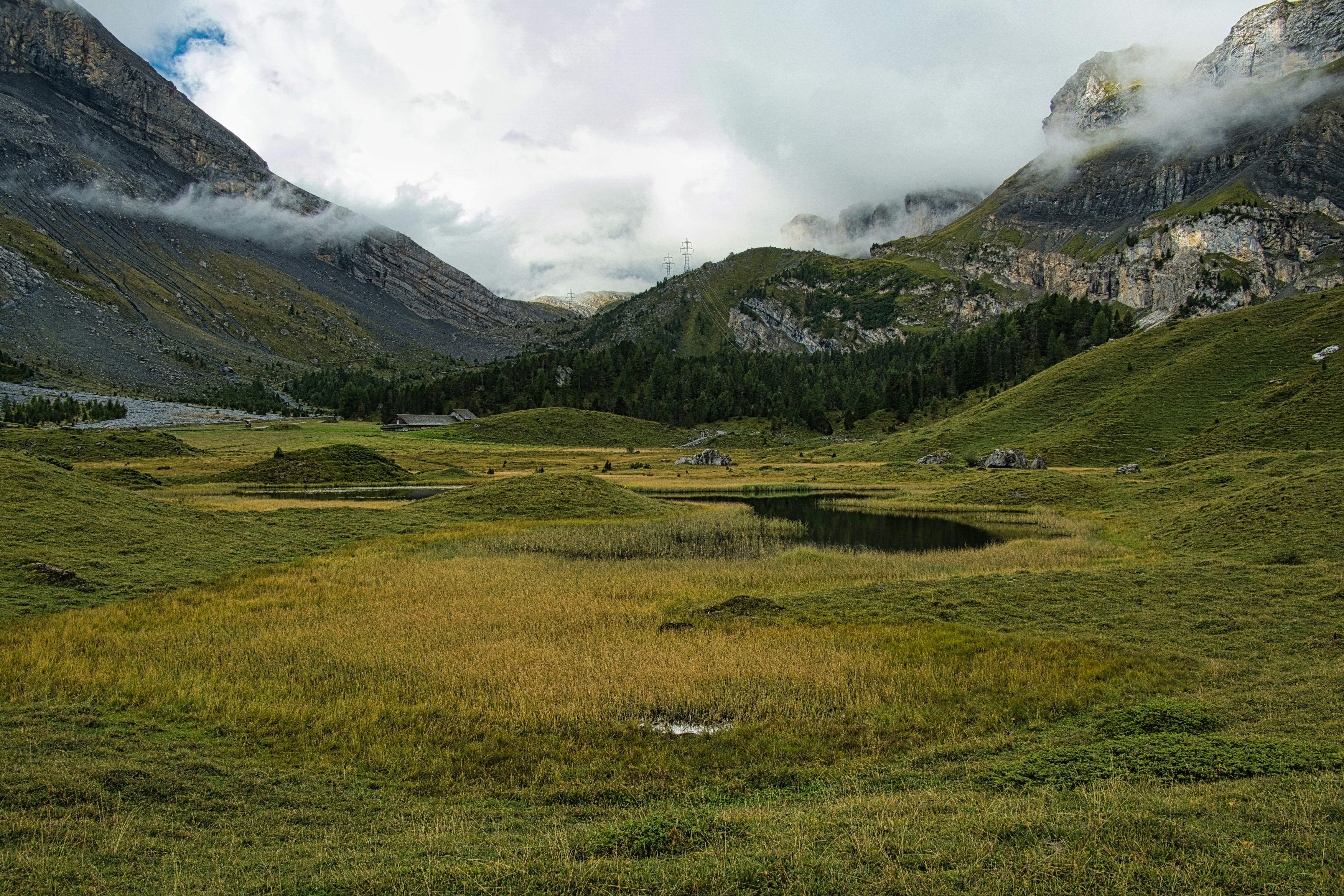 Soft alpine meadow with shallow ponds and distant mountains; low clouds drift over the mountaintops, creating a tranquil, expansive landscape.