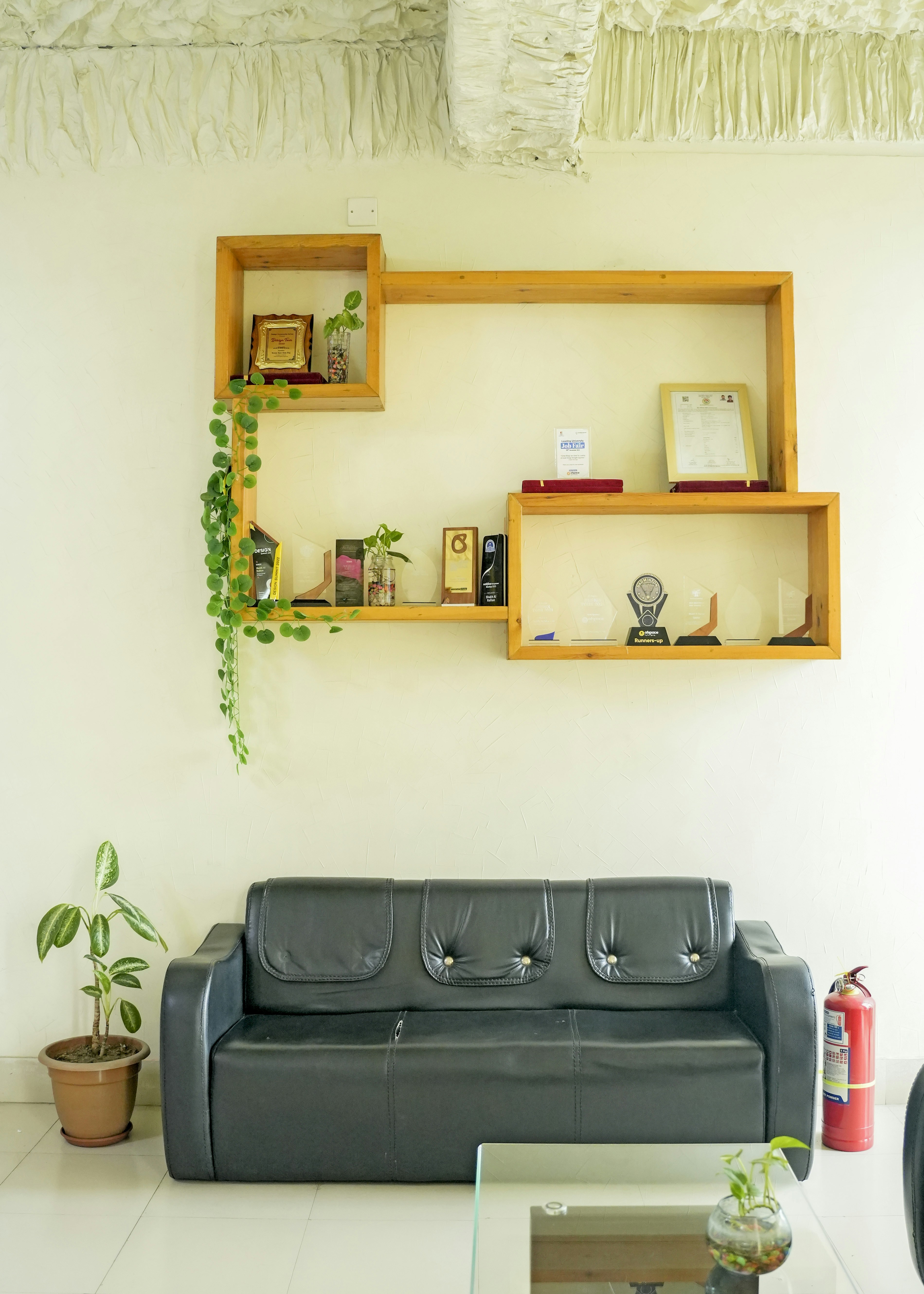 A tidy living room features a black leather sofa beneath a wooden staircase-style shelf arrangement and trailing green plant, with a fire extinguisher and small decor accents; a calm, contemporary space.