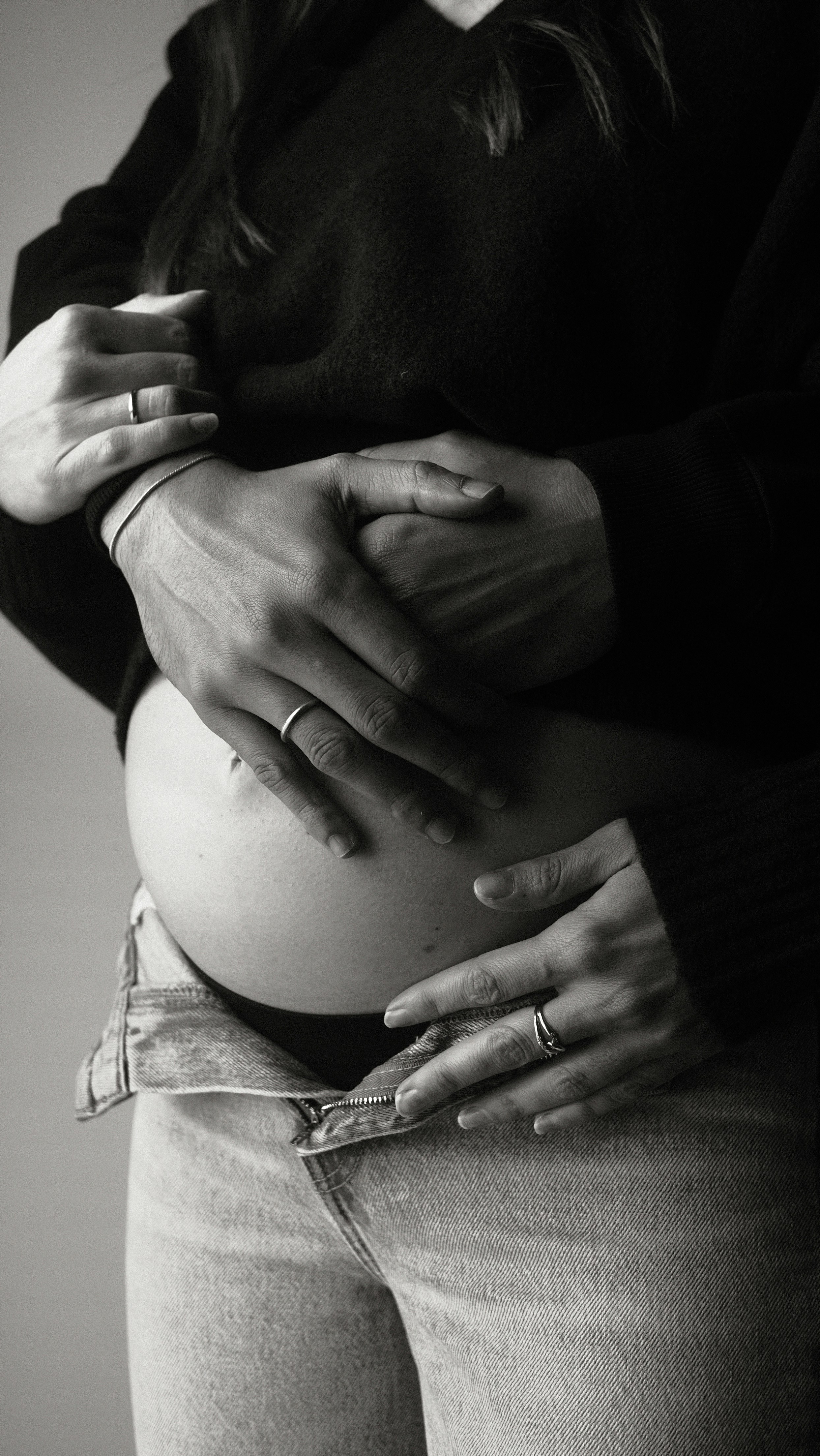 Close-up of a pregnant belly being cradled by multiple hands, wearing rings; monochrome portrait highlighting tenderness and anticipation.