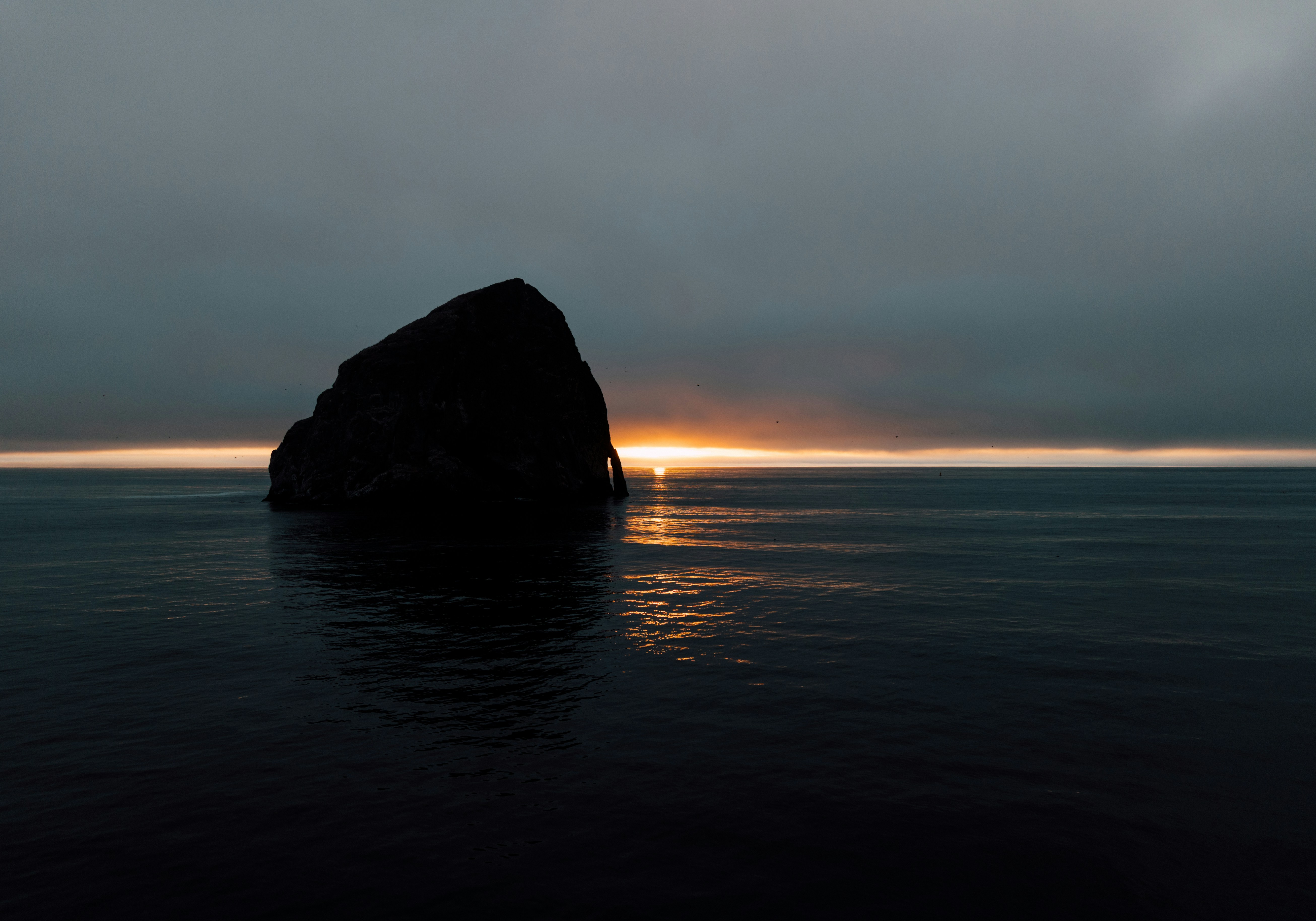 Dark sea stack rises from calm water as the sun peeks through a narrow gap in clouds along the horizon; the scene emphasizes subtle glow on the water and moody coastal atmosphere.