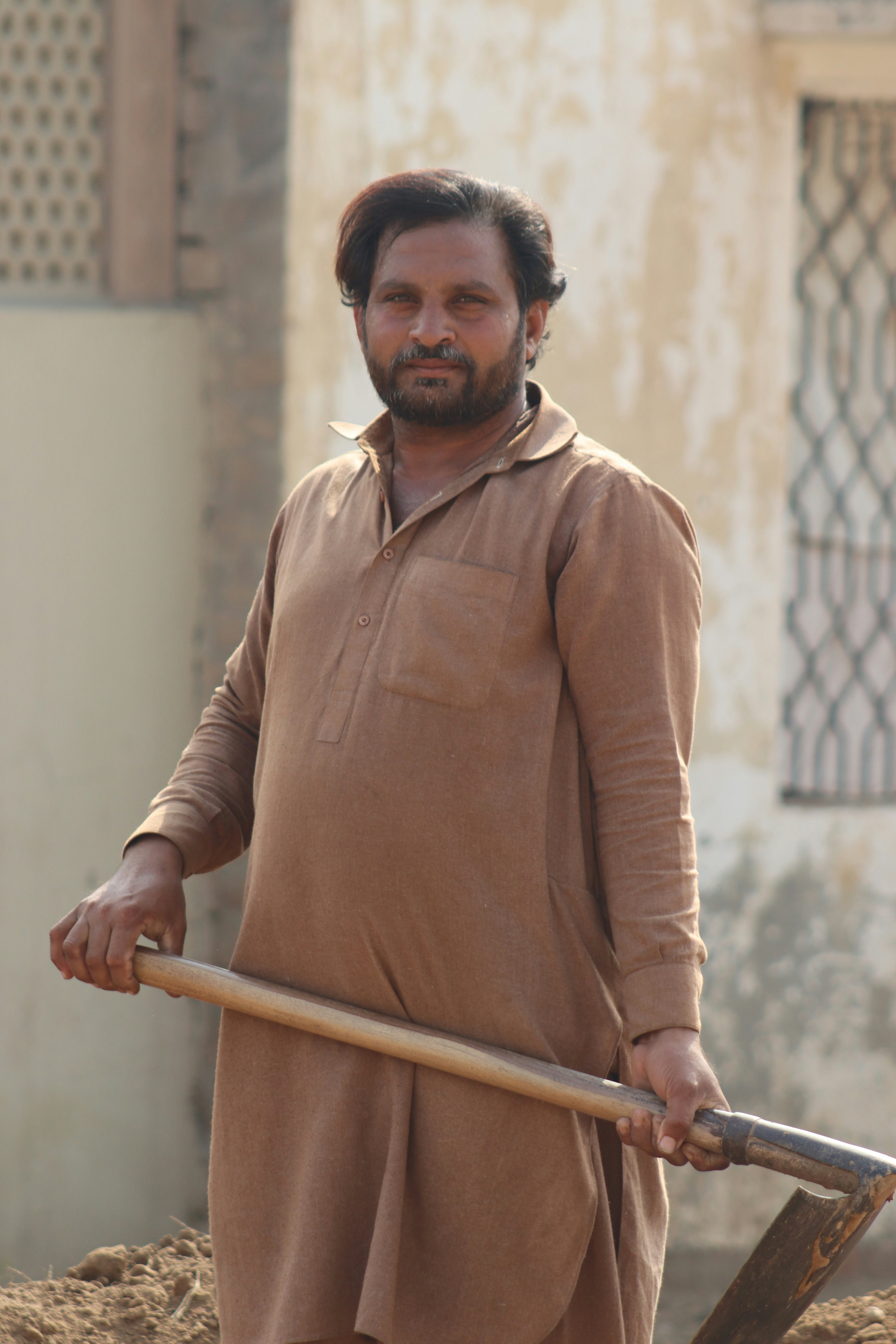 A man in a brown kurta stands outdoors with a shovel, against a weathered building. The scene focuses on his calm, steady posture and workwear in warm daylight. (photo is a candid portrait of a laborer with a tool, not a painting or logo).