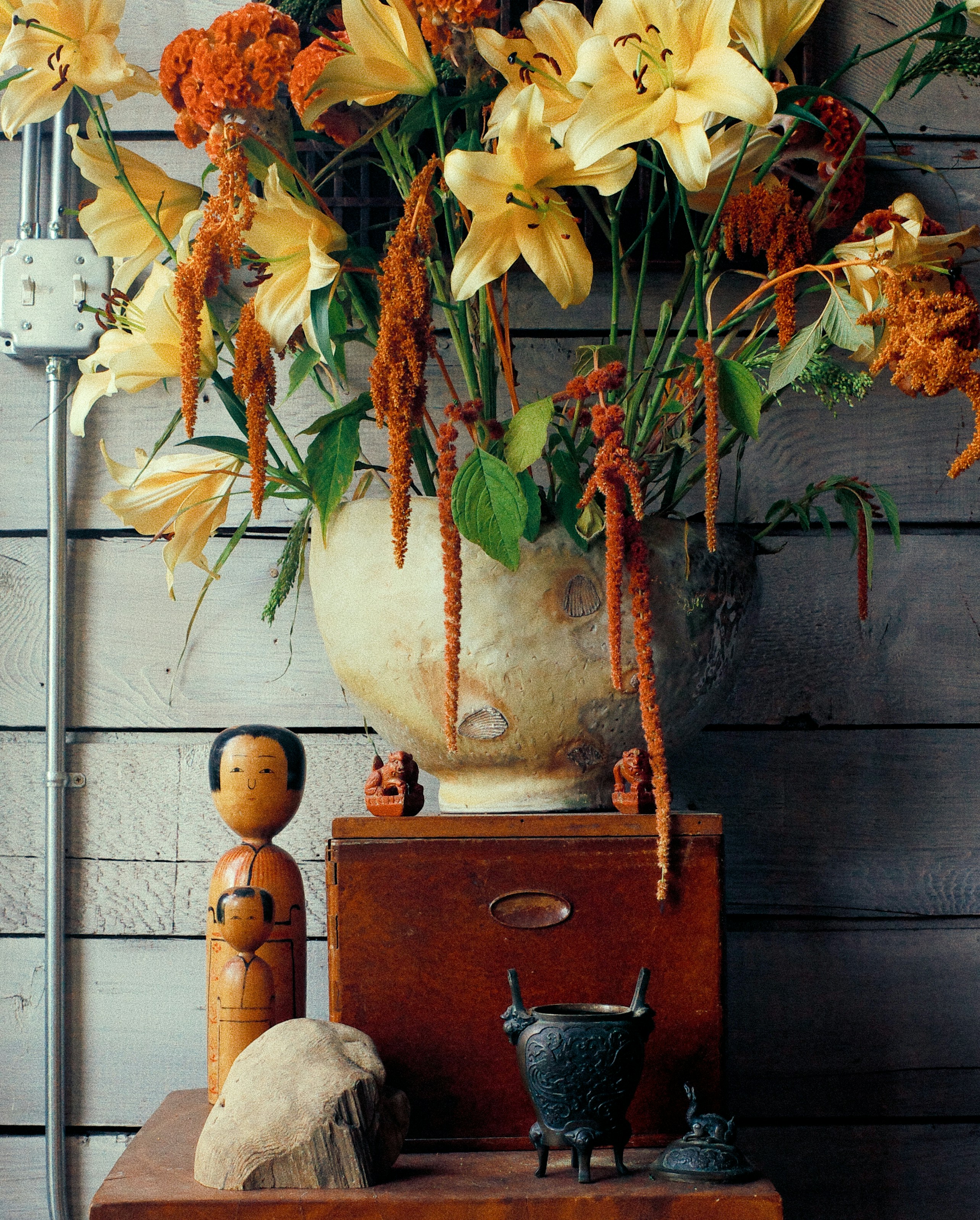 A still life arrangement featuring a large ceramic vase with yellow lilies and orange accents atop a wooden cabinet, flanked by carved wooden figures and small metal pieces against a weathered wooden wall.