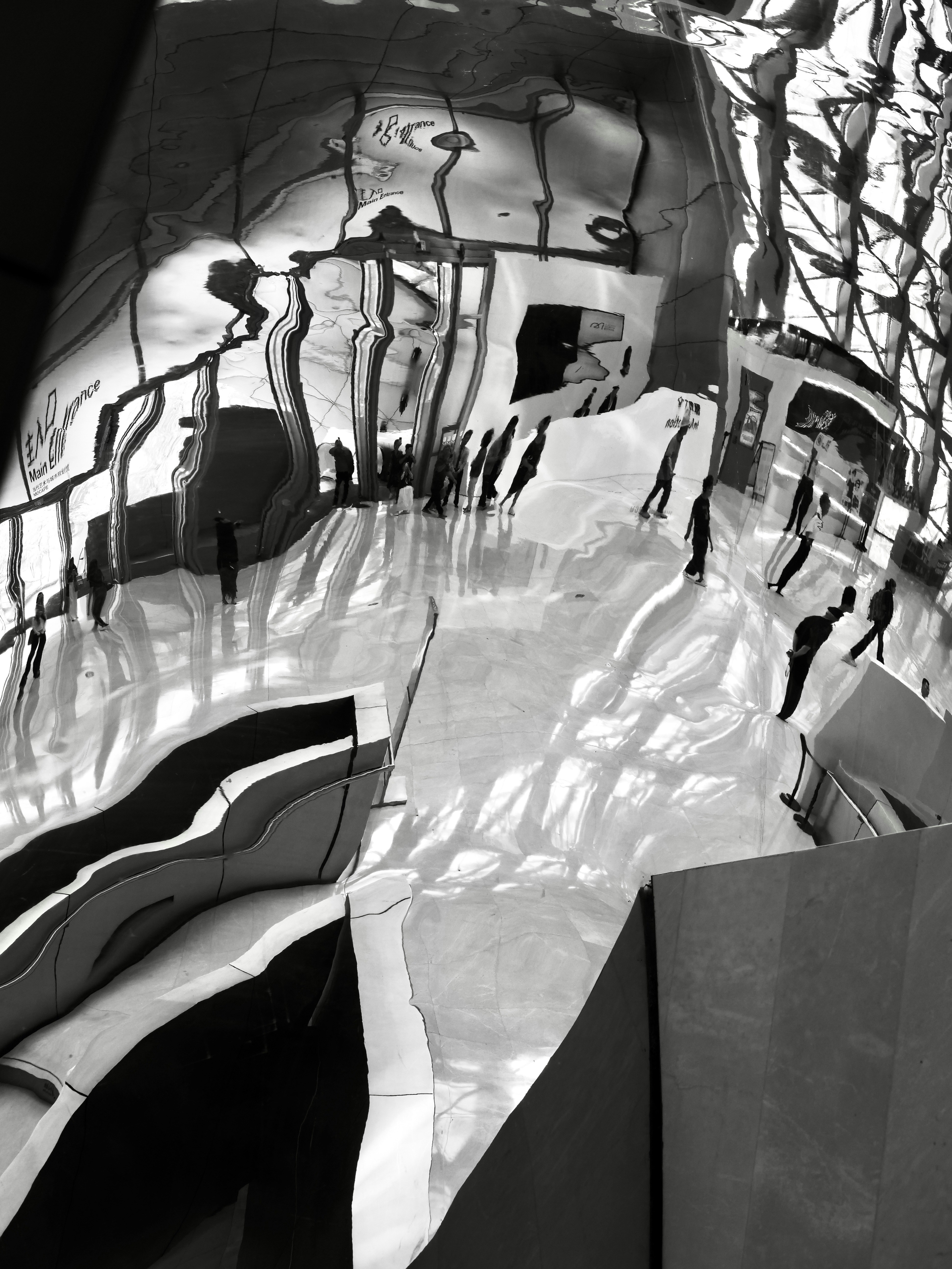 Black-and-white interior scene with distorted reflections of people walking in a glossy, sculptural space; a contemporary gallery or public installation with warped surfaces creating elongated silhouettes.