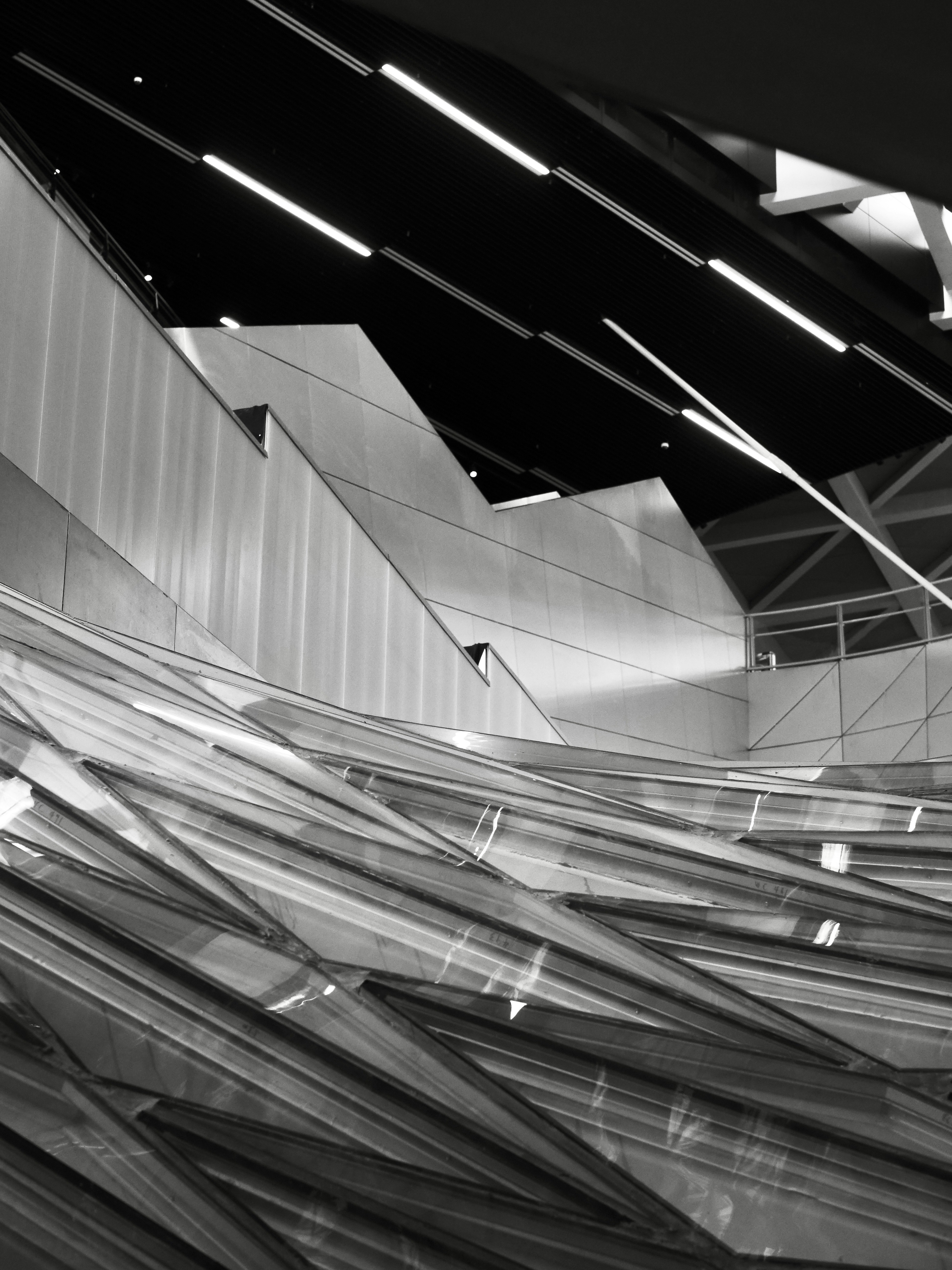 Monochrome architectural detail with diagonal glass and metal ramps creating dynamic ridges and lines across the frame. The composition emphasizes geometric textures and reflections.