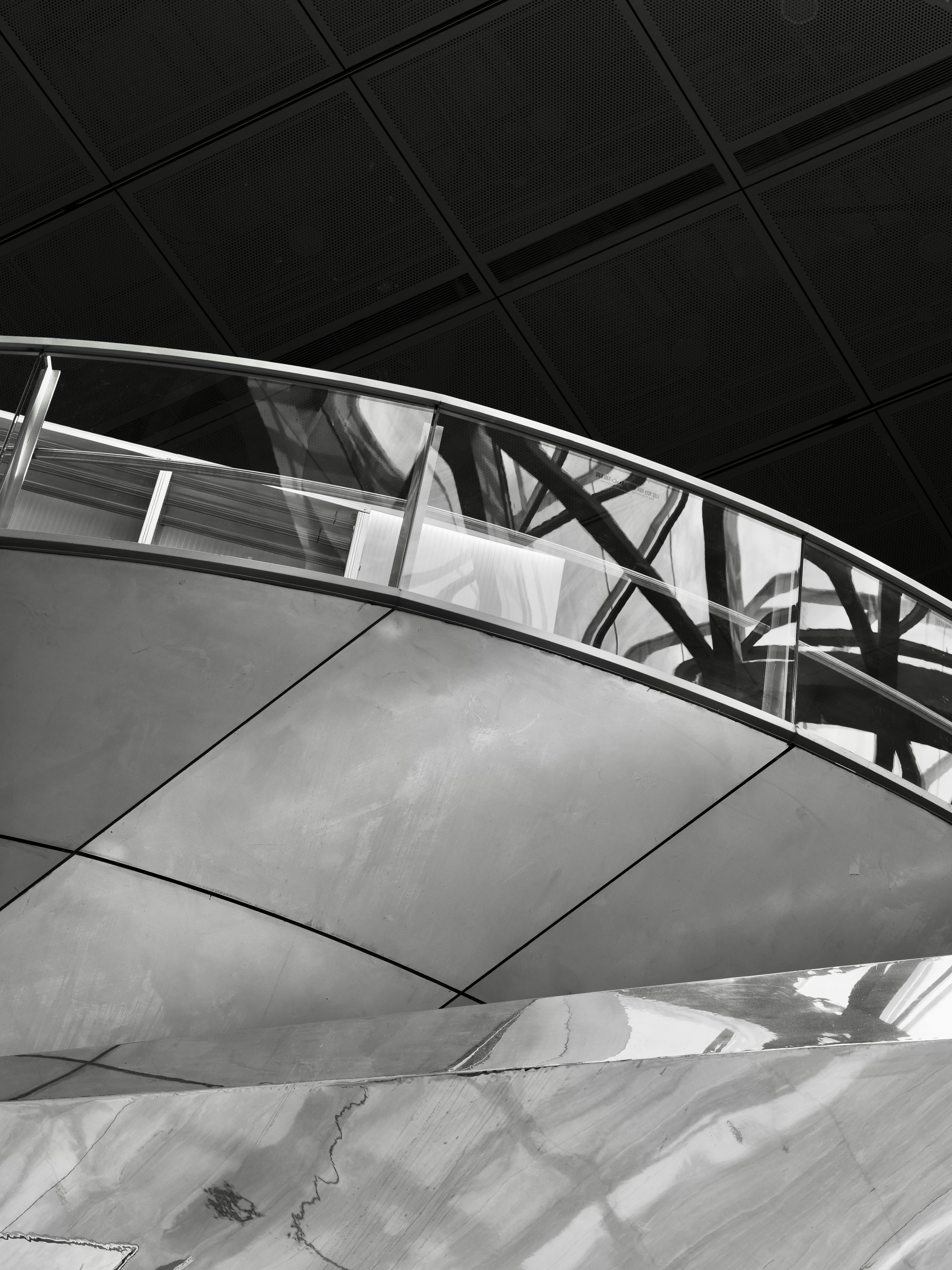 Monochrome abstract architecture featuring curved glass railing and reflective metal panels with strong diagonal lines and shadowed grid overhead.