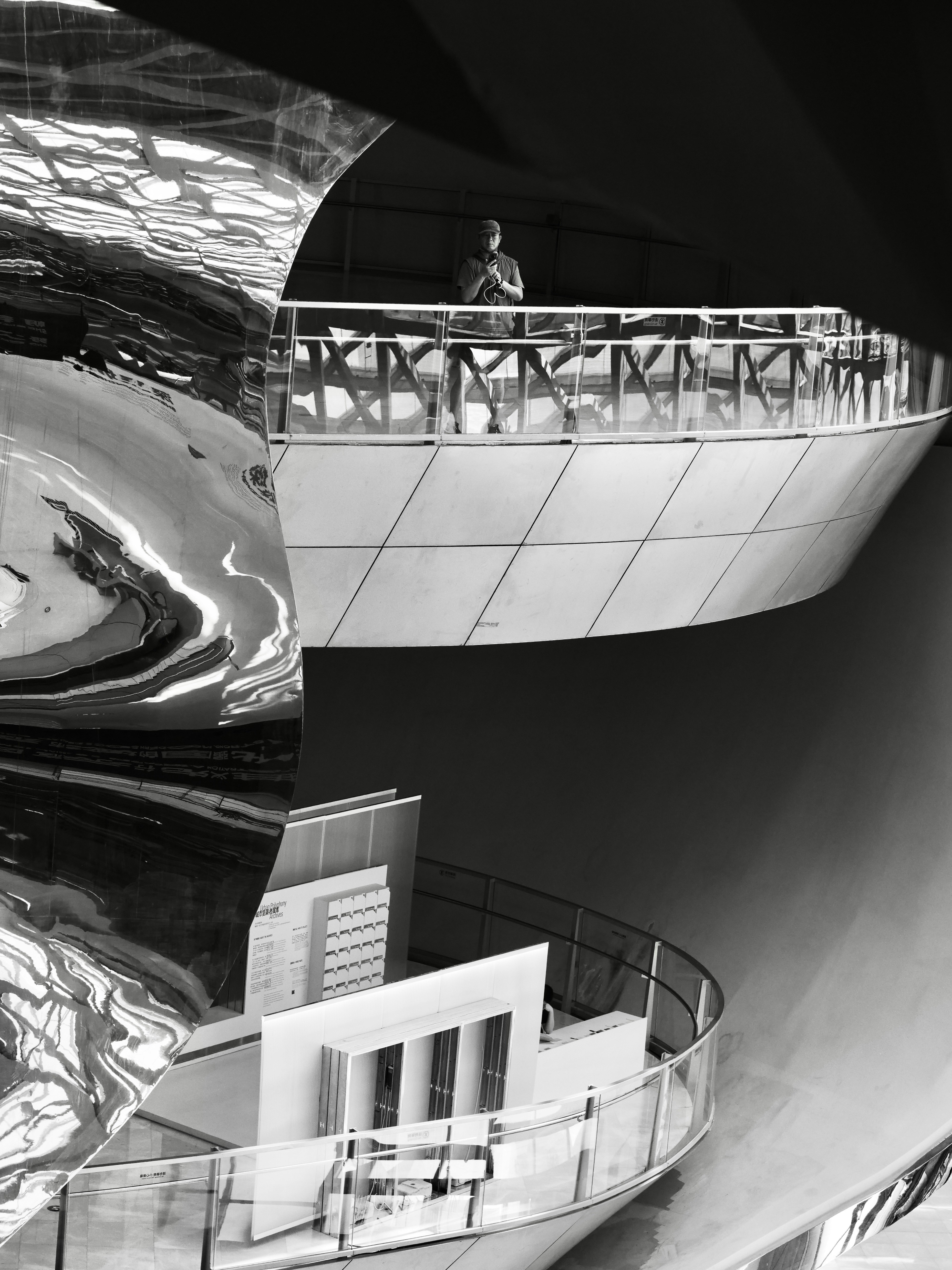 A monochrome interior with a suspended, curving walkway and a lone visitor leaning on the railing; architectural forms and reflections create a sculptural composition.