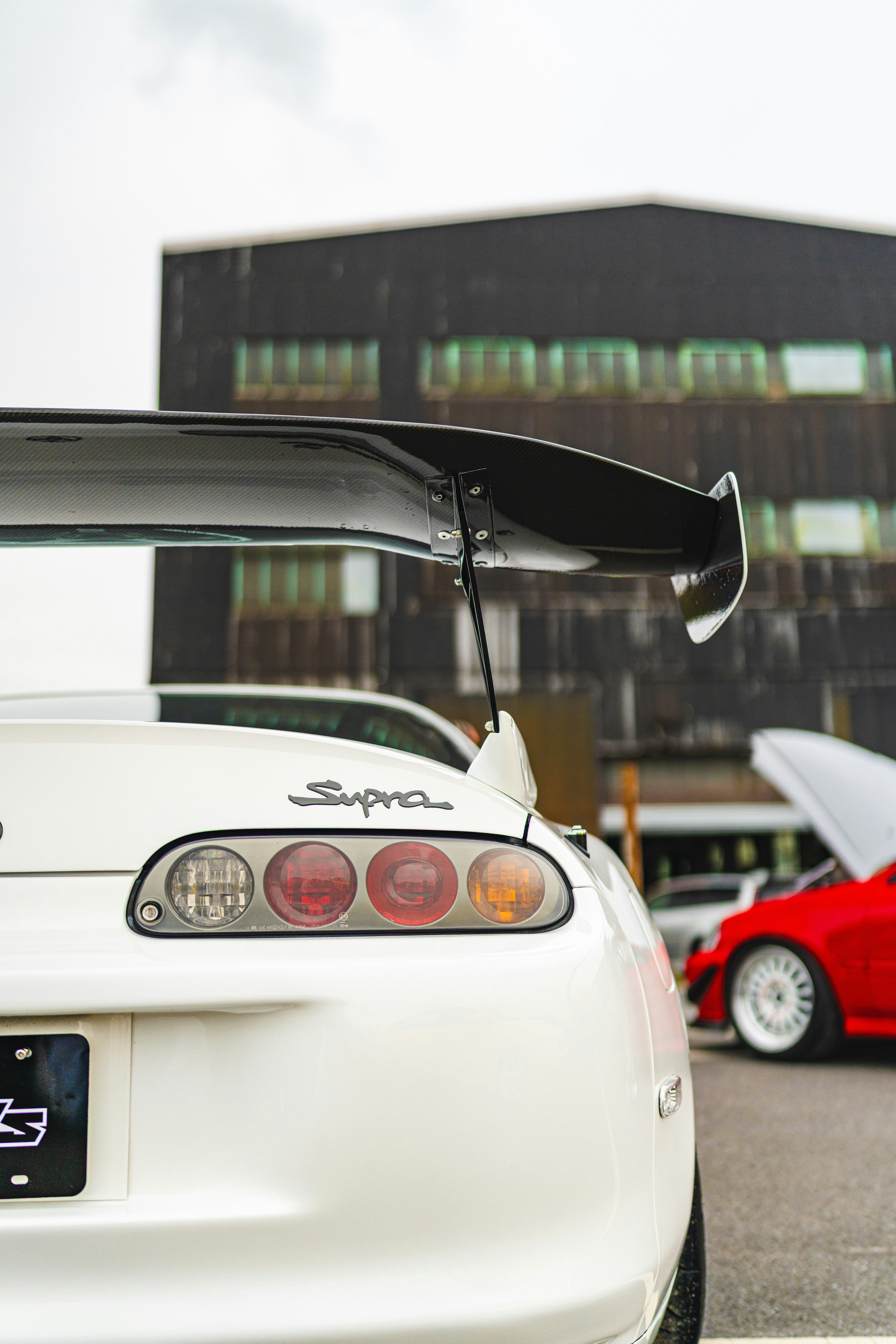 White sports car with large spoiler parked in front of a dark industrial building; tail lights and ' Supra' badge visible, with a red car in the background.