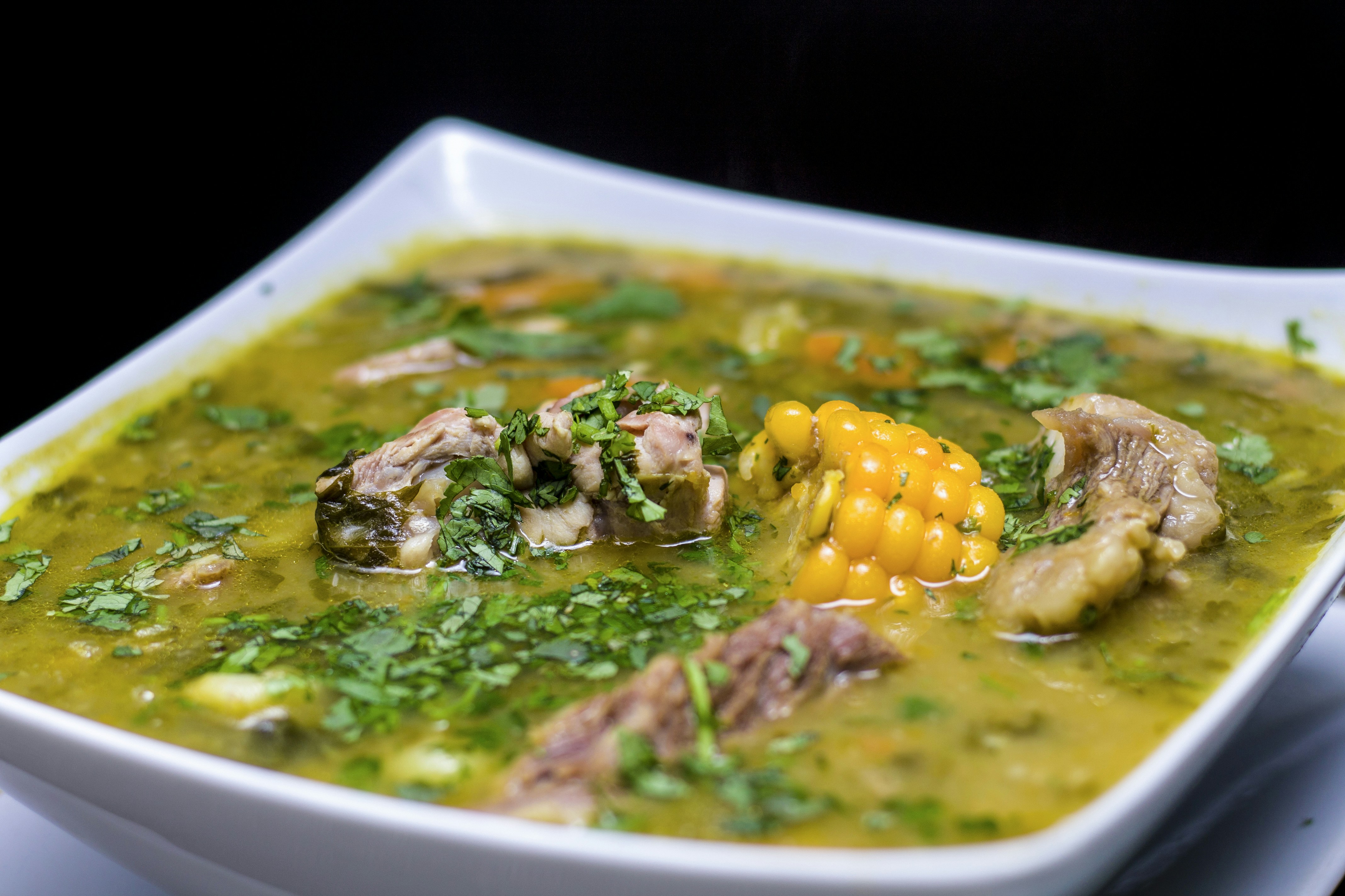 Creamy green soup with chunks of meat and a bright corn cob, garnished with fresh herbs in a white bowl.
