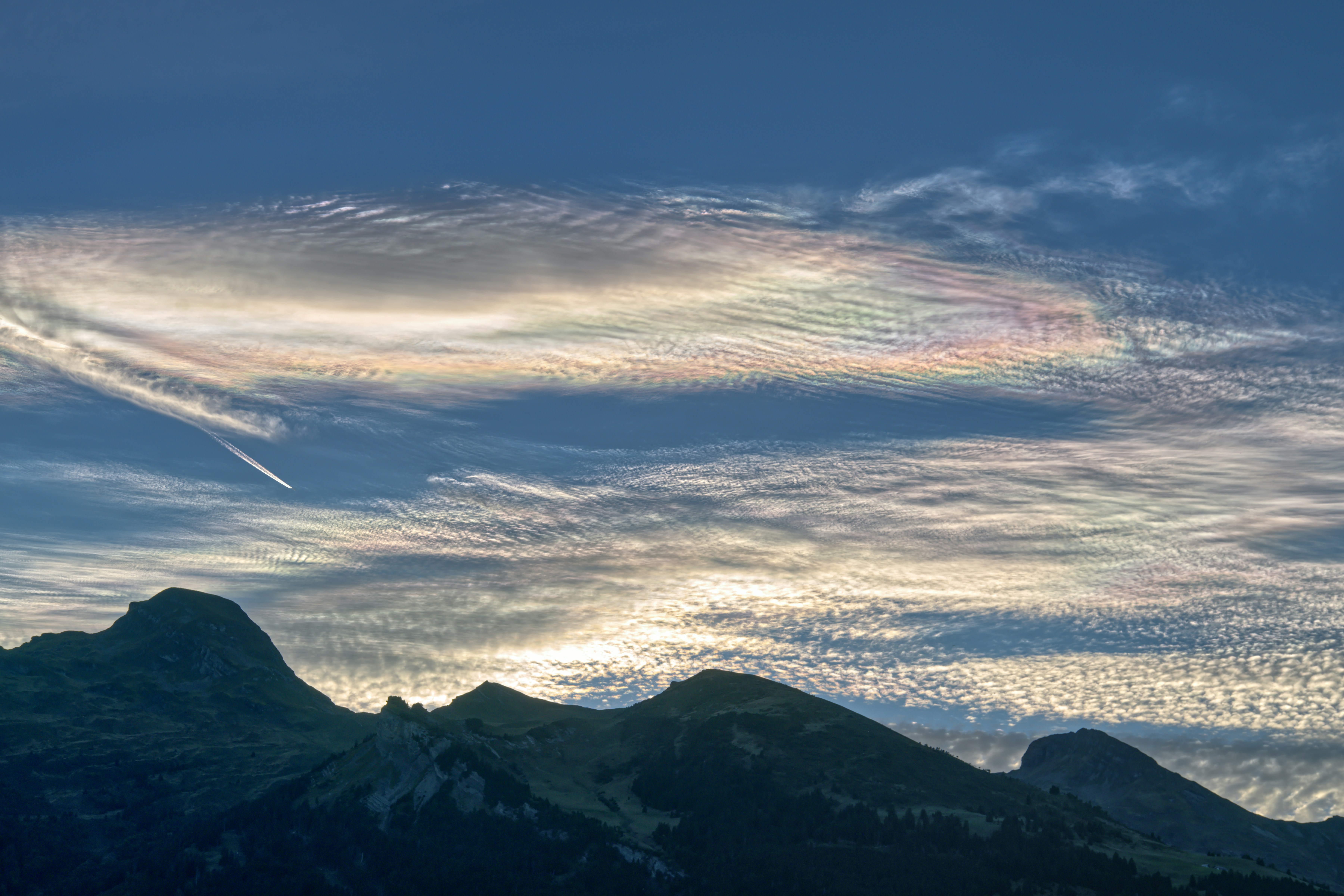 Distant mountain silhouette under a dramatic, cloud-washed sky with a contrail streaking diagonally across the left. A calm landscape emphasizing the horizon and atmospheric color gradations.