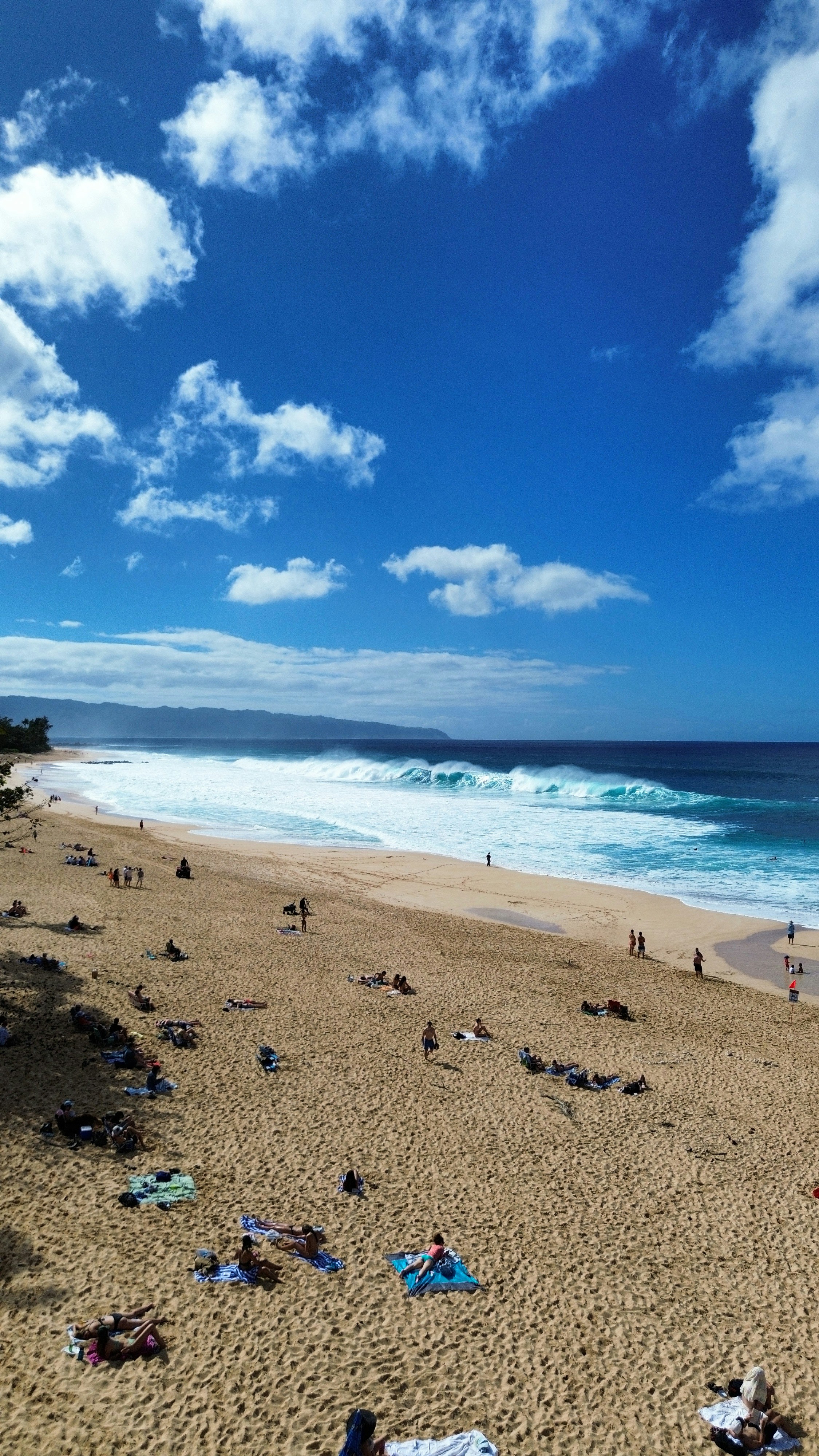 Vast beach scene with blue sky, white clouds, and foamy waves; people scattered across sandy shore with towels and umbrellas. Aerial view shows horizon where ocean meets coastline; the setting emphasizes leisure activity and coastline.