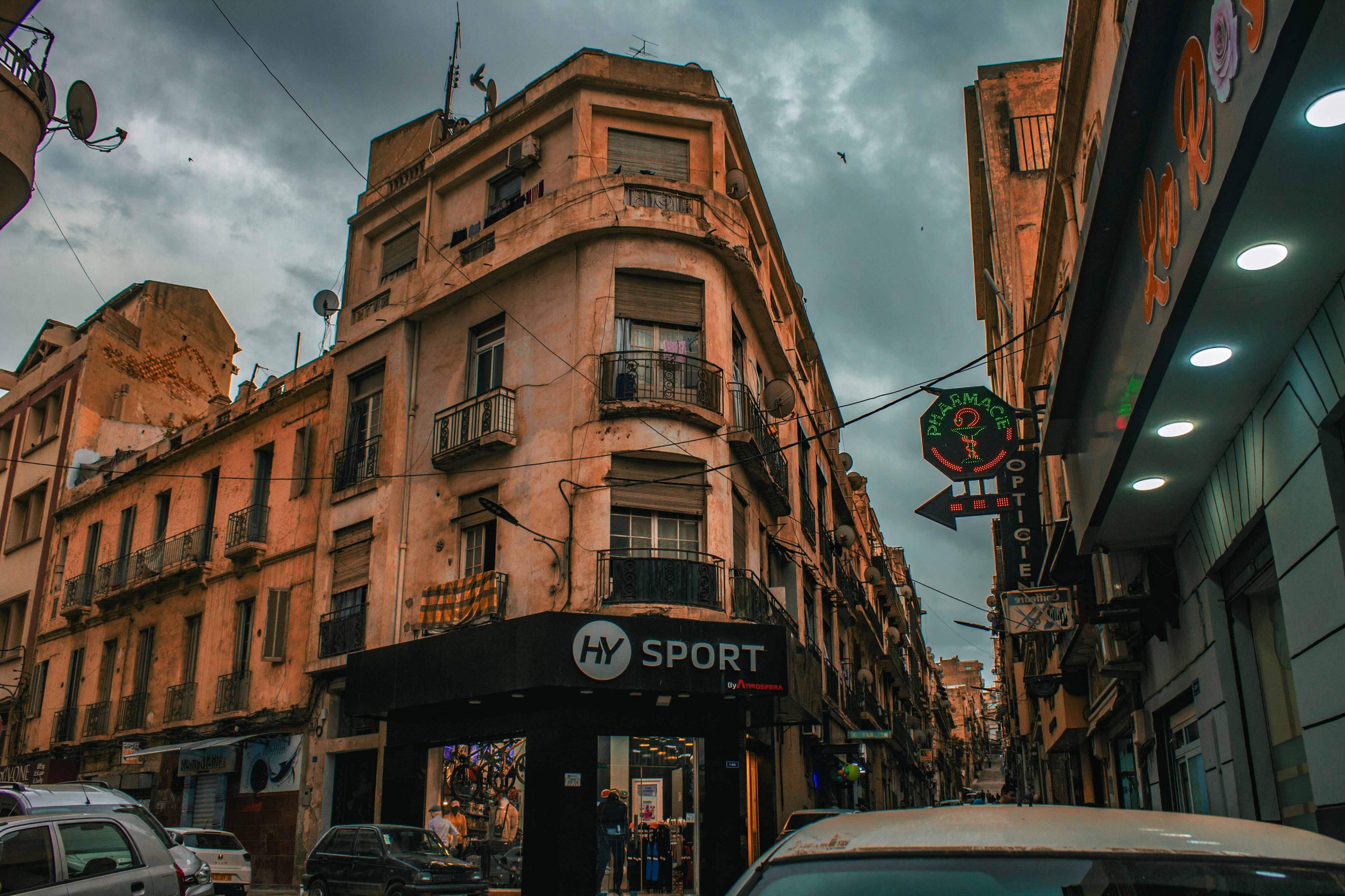 A bustling city street with aged buildings lining the sidewalk and a neon-lit shopfront; parked cars frame the foreground, conveying a late-day urban atmosphere.