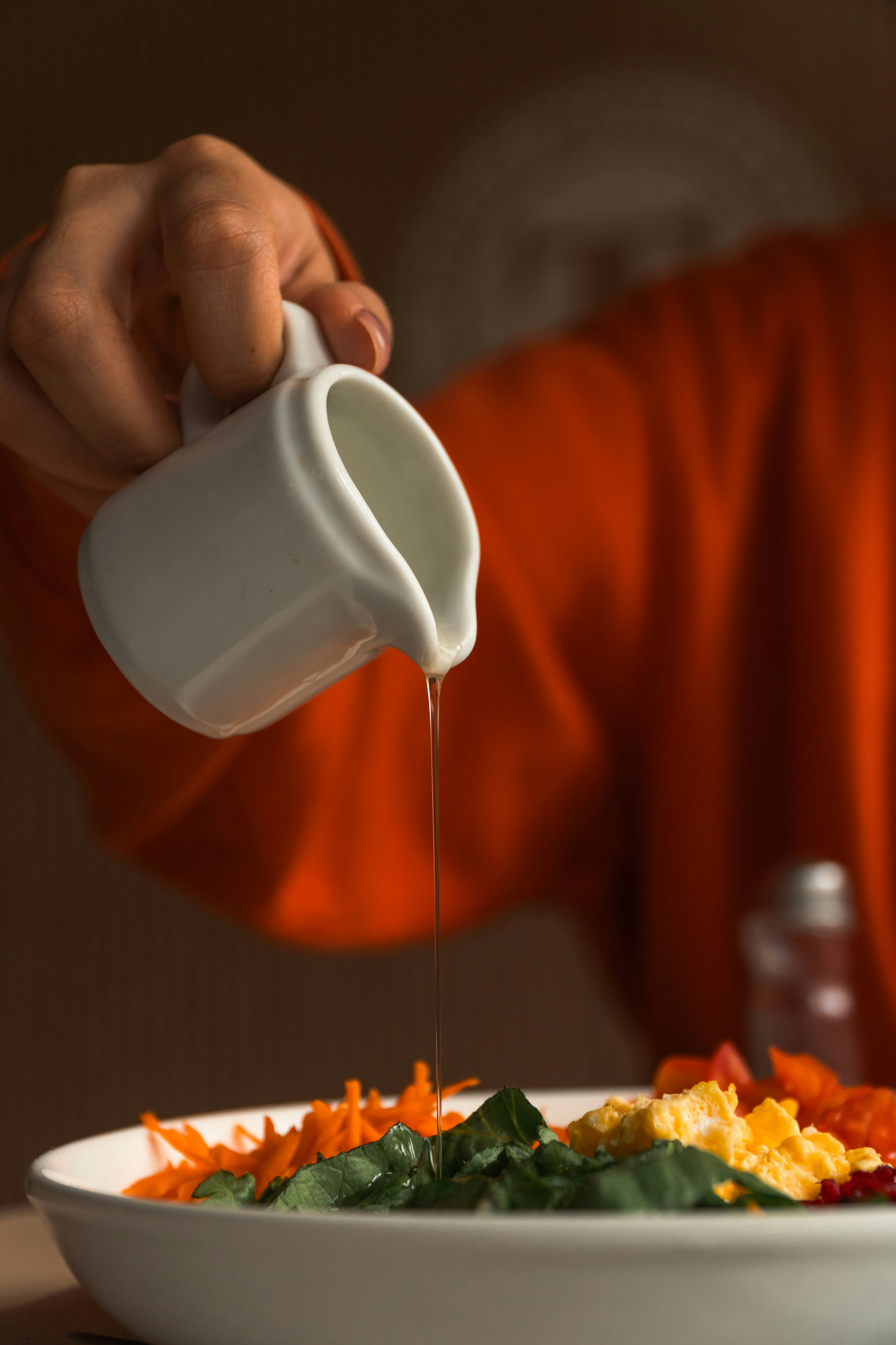 A hand tilts a small white jug, gracefully pouring oil over a colorful salad in a shallow bowl. The scene emphasizes texture and depth, captured in warm, intimate lighting.