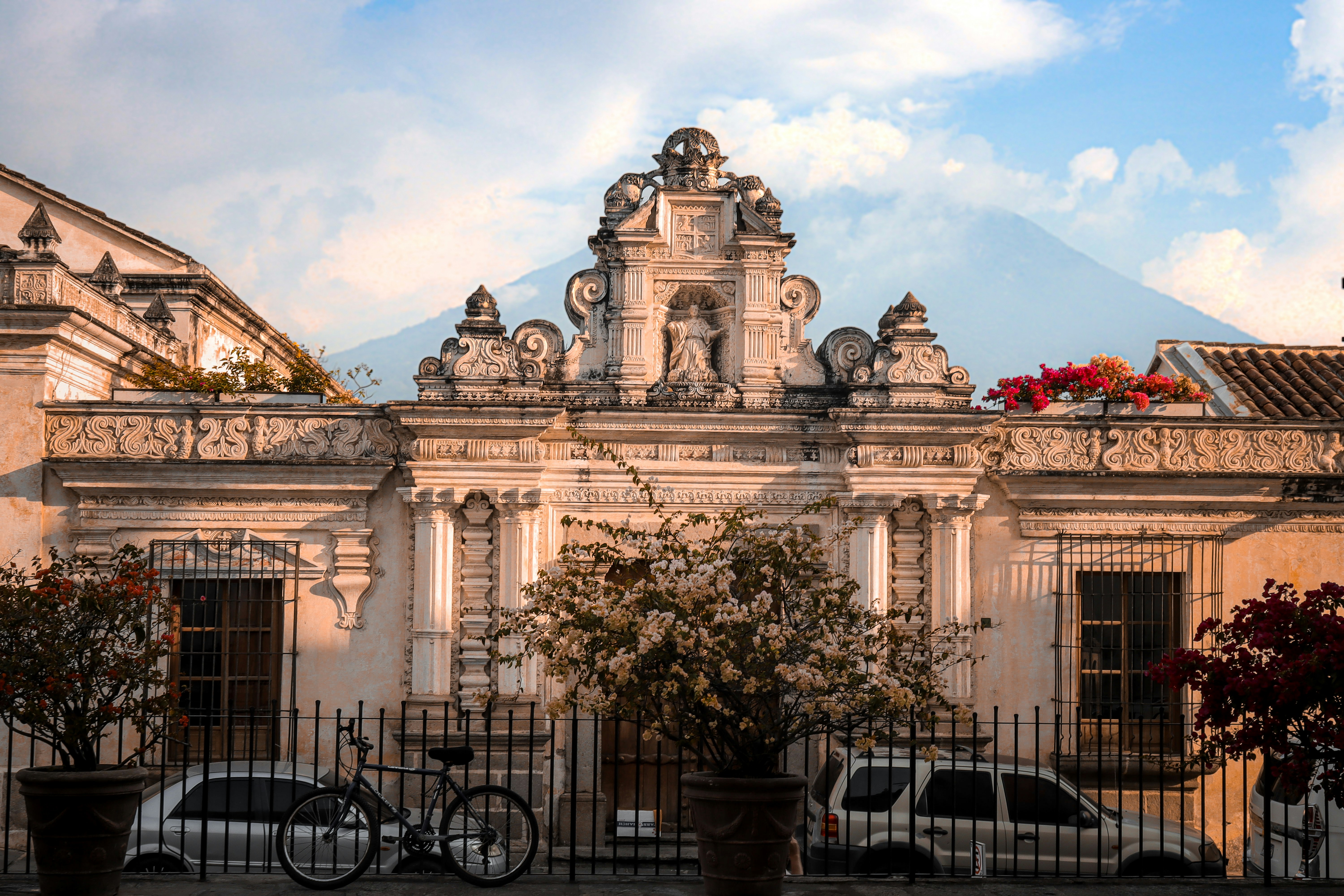 Sunlit colonial facade with intricate architectural details and blooming trees in the foreground; motorcycles and bicycle lean against a low fence, mountain peak visible in the distance. A logo-like central building anchors the scene.