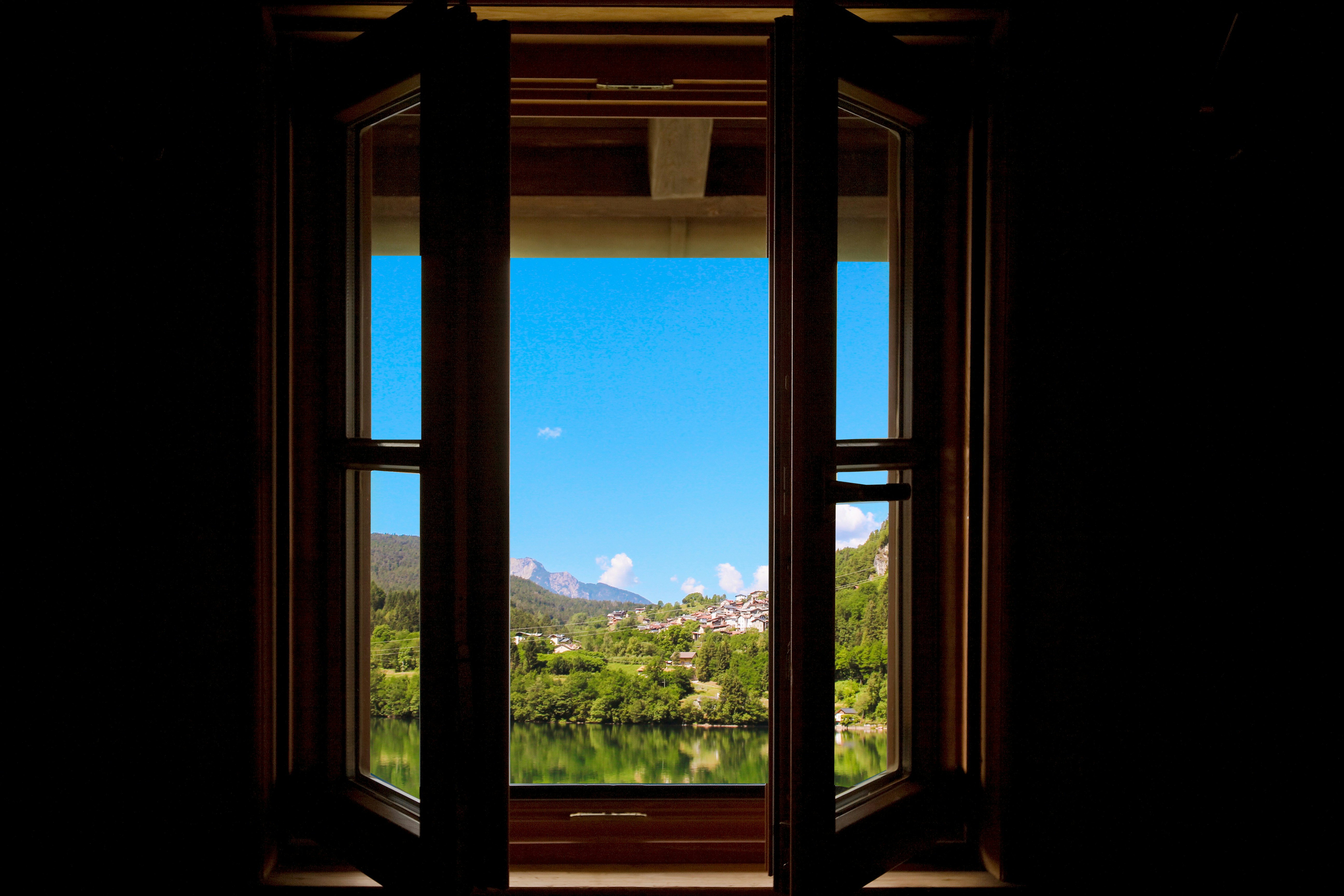 A bright landscape view is framed by an open window, revealing blue sky, distant hills, and a calm lake; the scene emphasizes contrast between dark interior and vivid exterior. This is a photographic capture of a window scene.