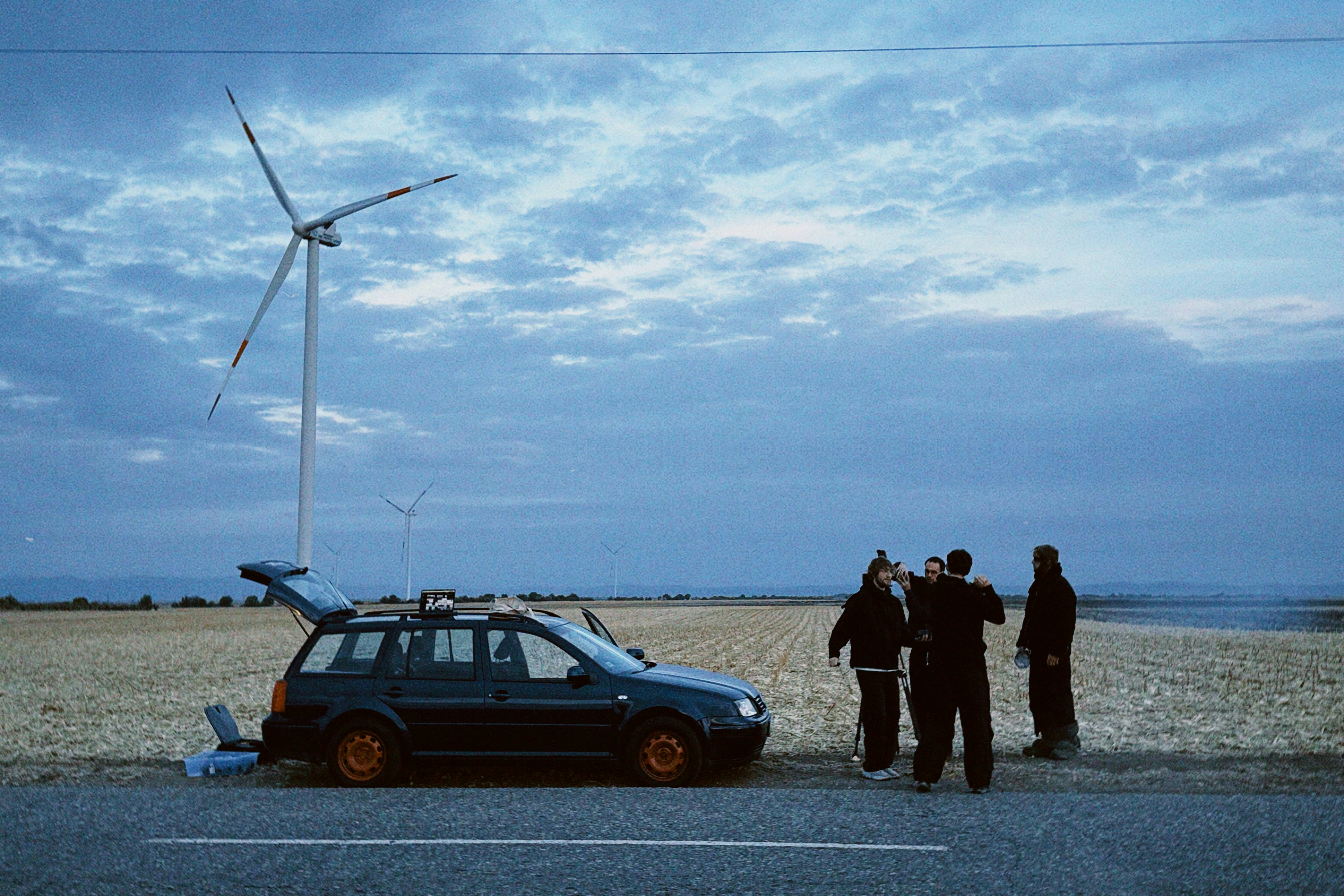 Group of people stands near a car on a quiet coastal road with a wind turbine in the background, suggesting a roadside gathering at dusk near open beach.