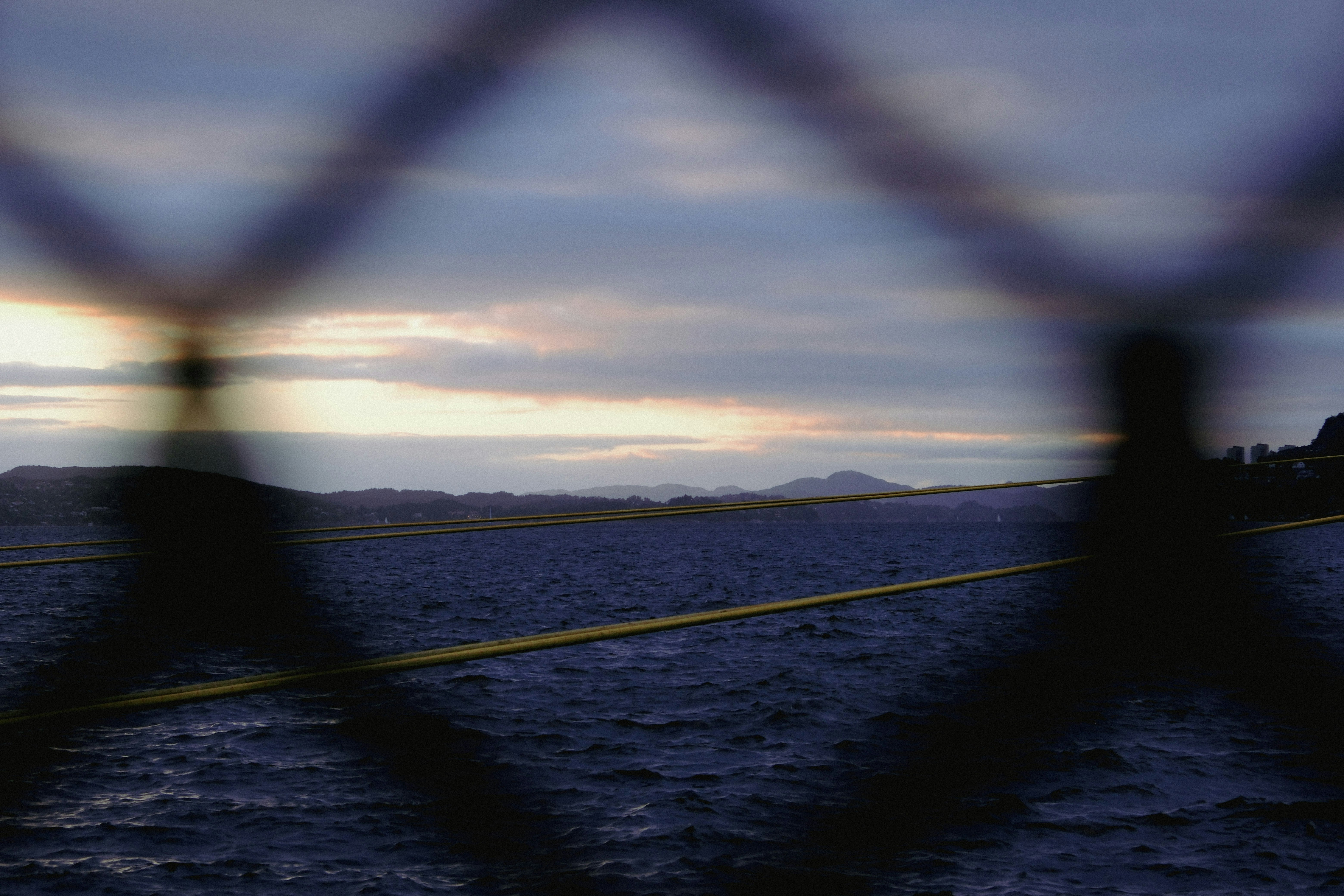 Distant coastline and sunset behind a blurred chain-link fence, creating a moody urban-seascape with a sense of barrier and distance.